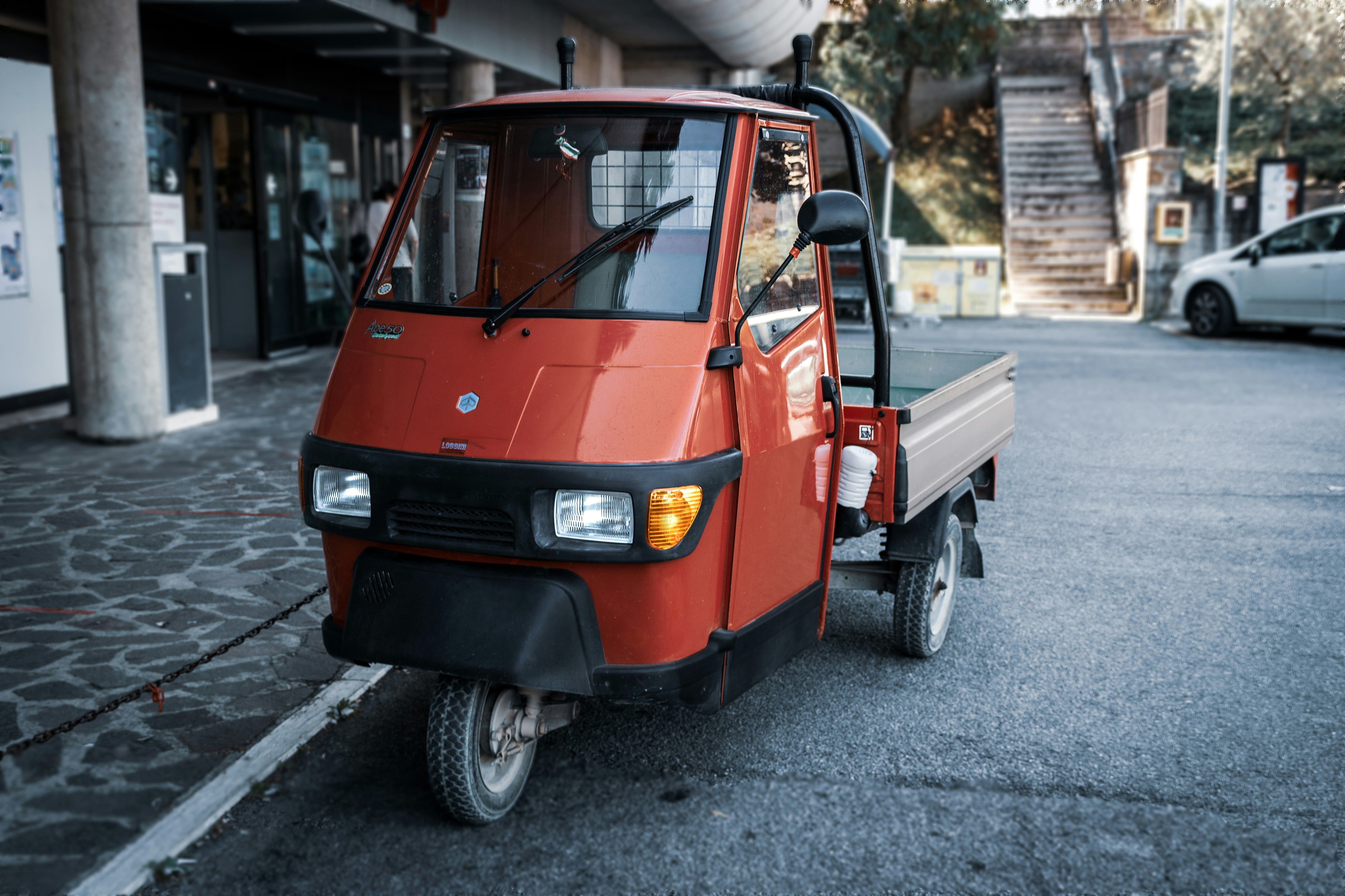 A vibrant red three-wheeled vehicle parked on a city street, showcasing its compact design and utility. The background features urban elements, enhancing the scene's character.