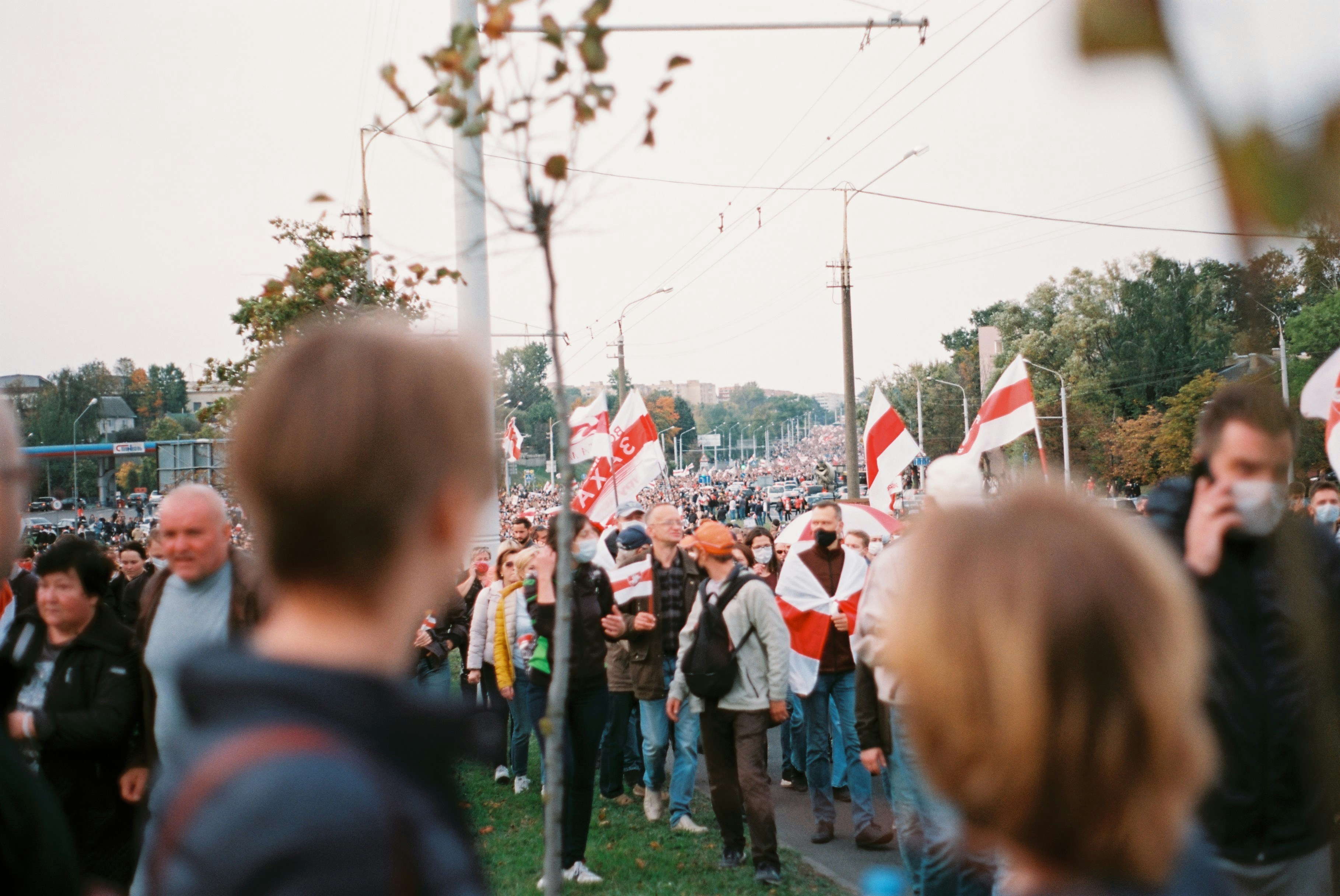 A street crowd waves red-and-white flags while foreground figures are blurred, and the mid-ground crowd remains legible, conveying a public gathering with motion and focus on the flags.