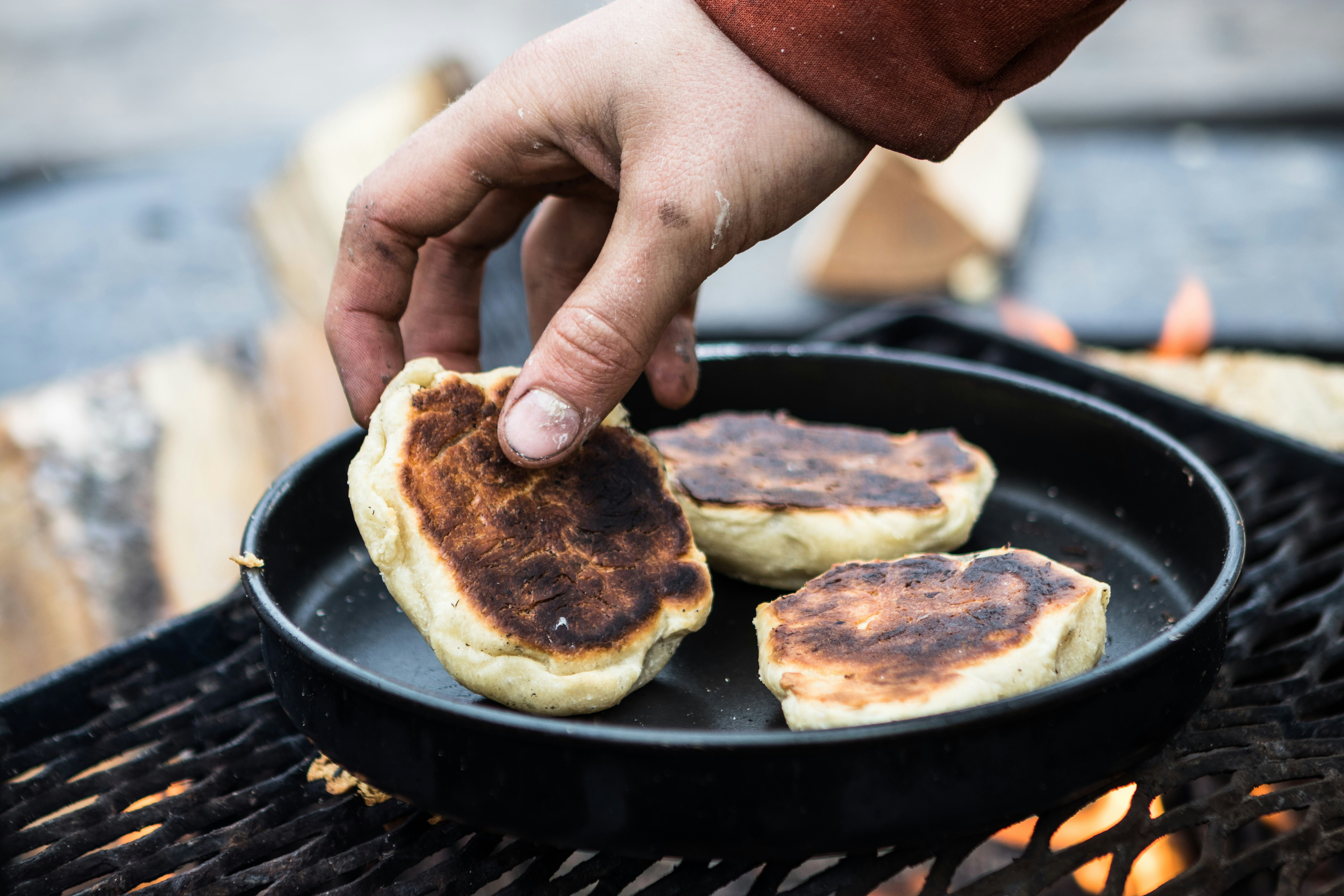 Hand reaching for golden-brown baked goods resting in a pan over an open fire, showcasing outdoor cooking techniques.