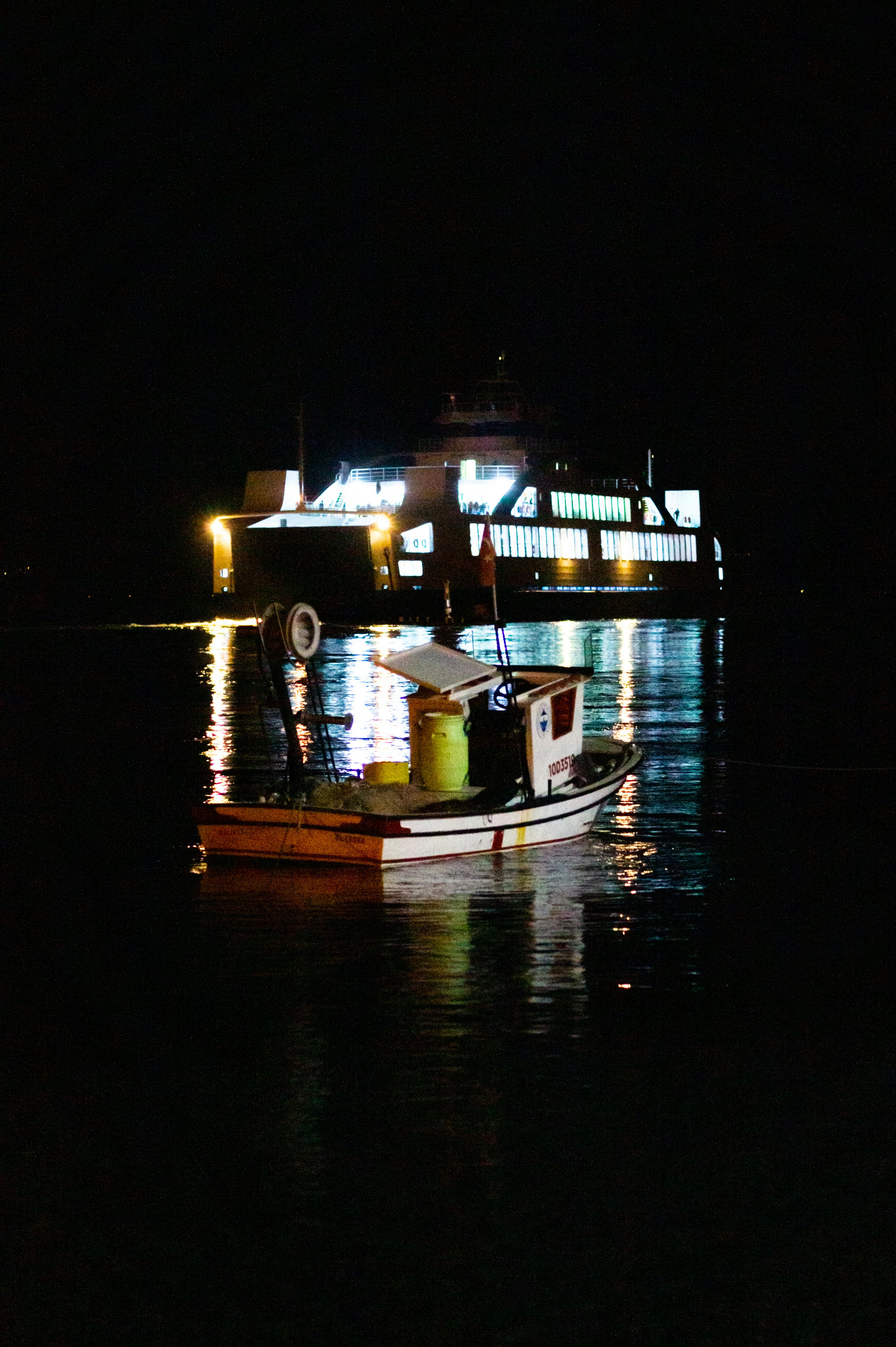 A small fishing boat anchored in a calm harbor, illuminated by the distant glow of a ferry's lights reflecting on the water's surface.
