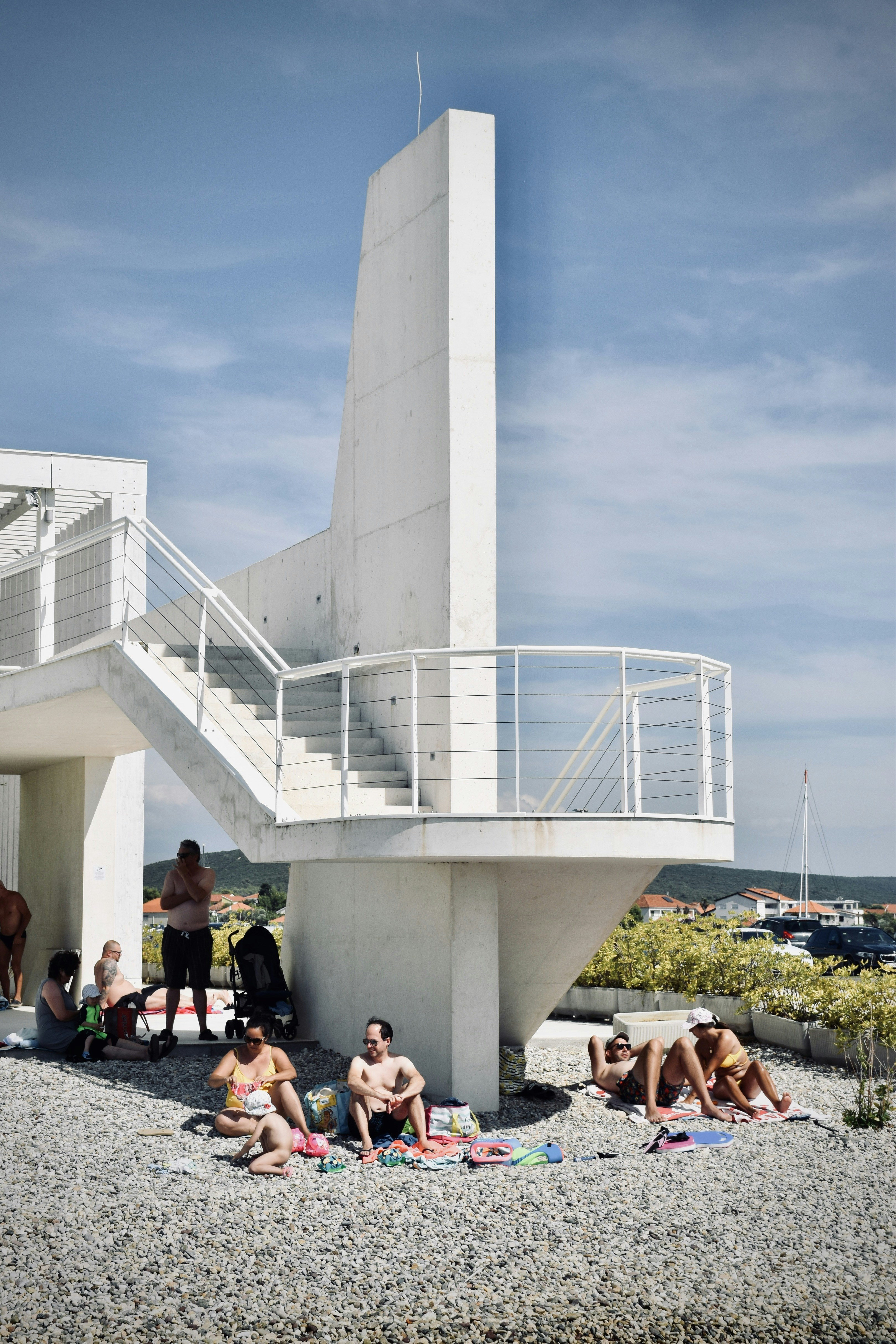 Beachgoers relax on pebbles beneath a striking architectural structure, enjoying a sunny day by the water's edge.