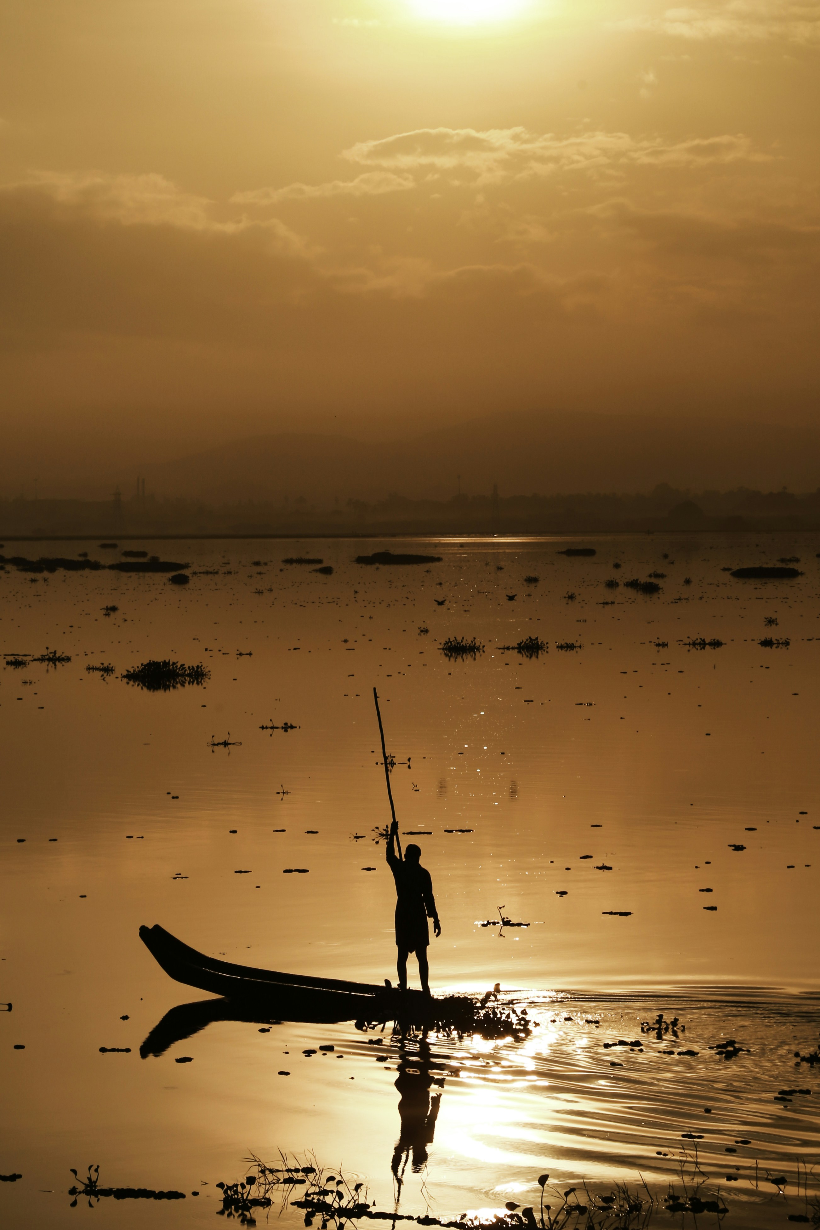 A lone fisherman stands in a boat, silhouetted against a golden sunset over a tranquil lake, with gentle ripples reflecting the fading light.