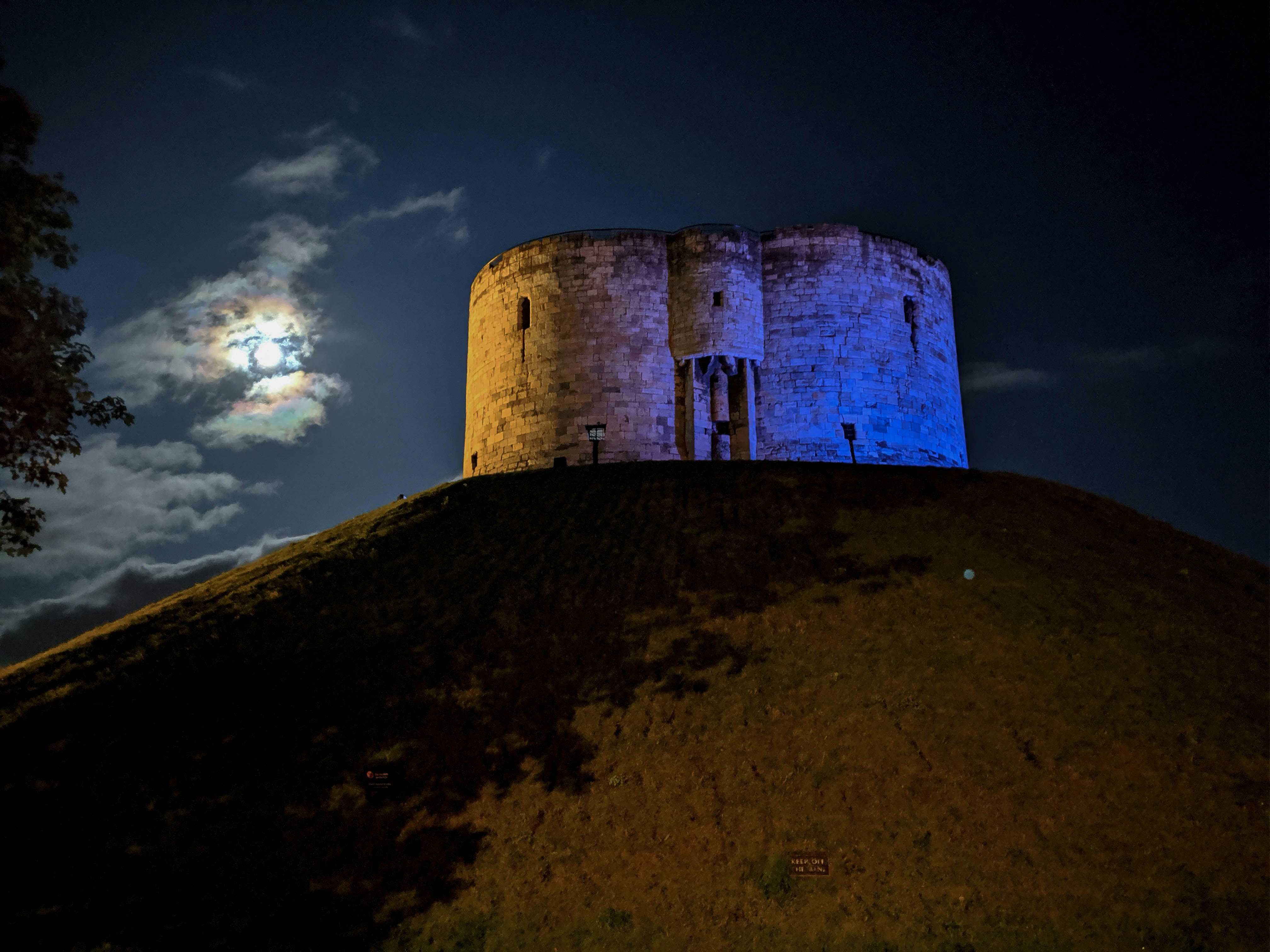 Historic stone tower illuminated by blue light under a full moon, surrounded by a night sky with scattered clouds.