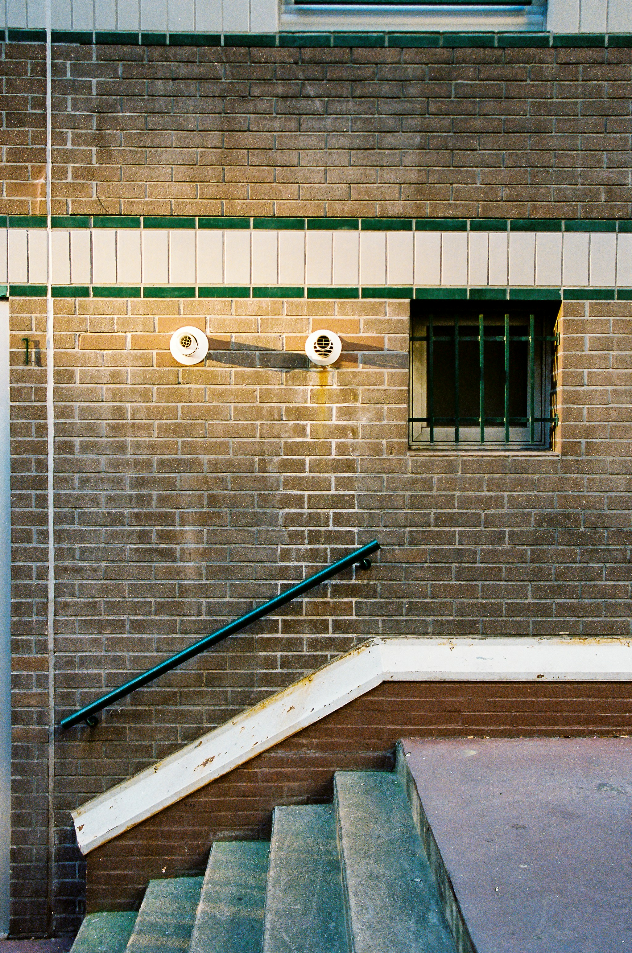Brick wall with a diagonal green handrail, a barred window, and two circular vents create a texture-forward architectural composition.