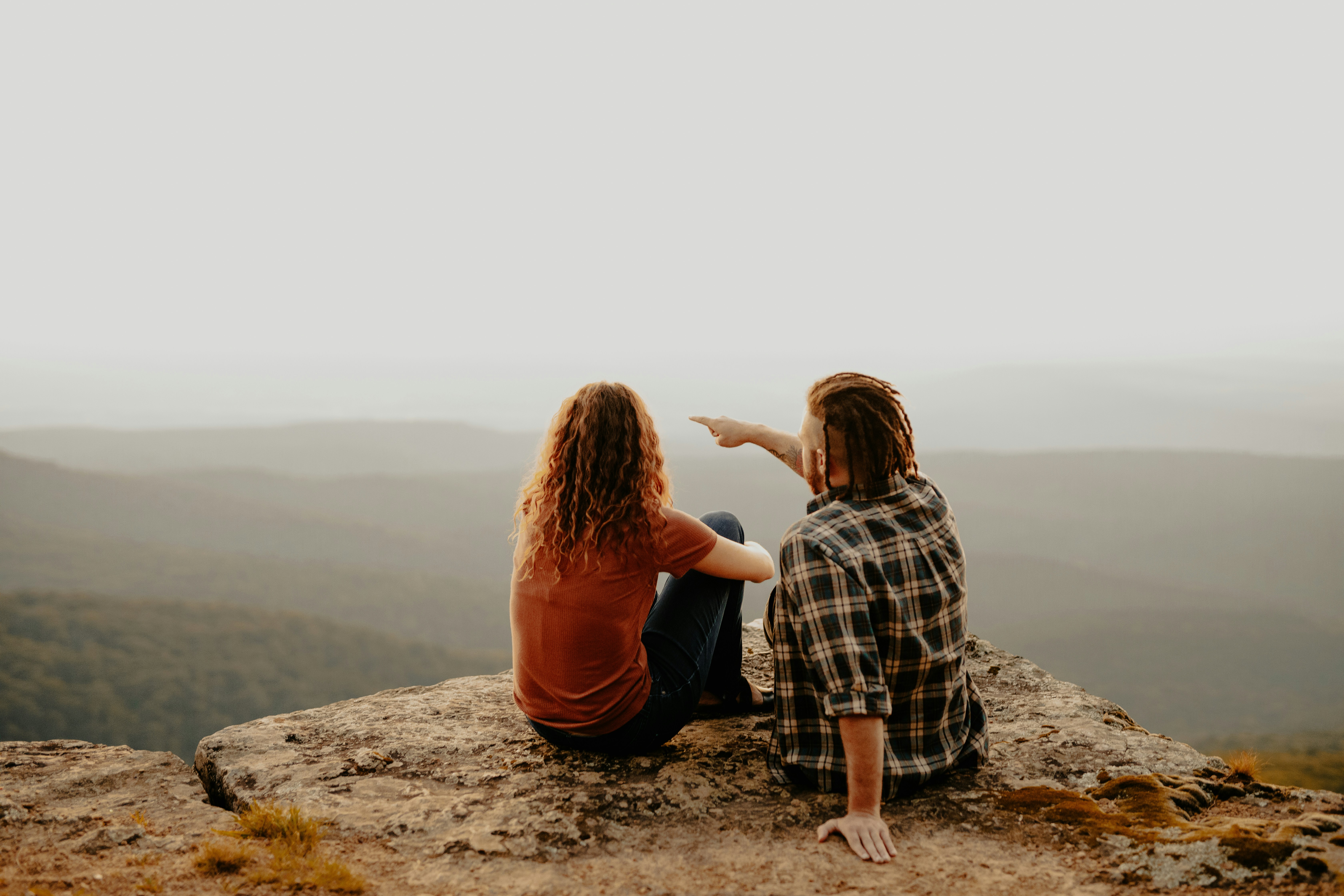 Two individuals seated on a rocky ledge, engaged in conversation while overlooking a vast, misty landscape.