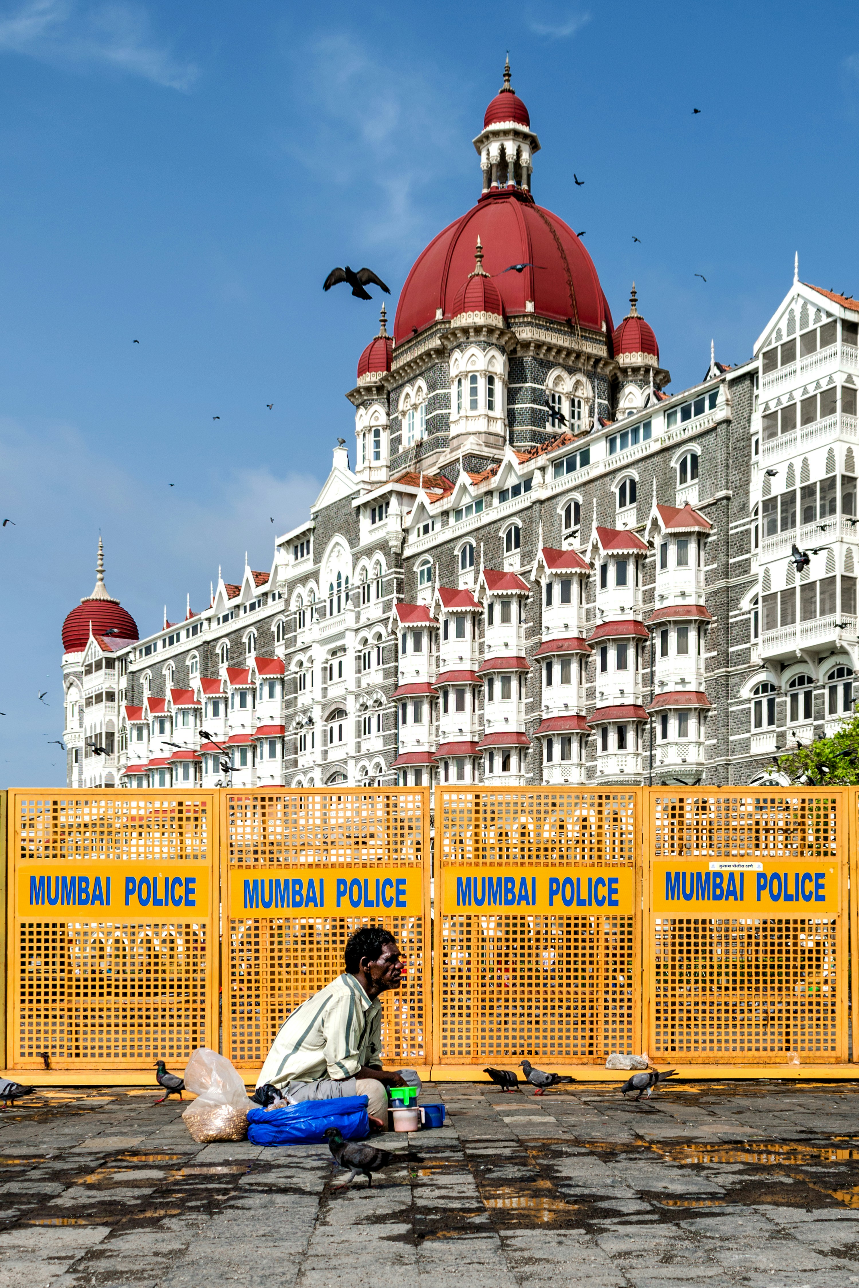 A street vendor sits against a backdrop of the iconic Taj Mahal Palace hotel, surrounded by police barricades and birds in flight.