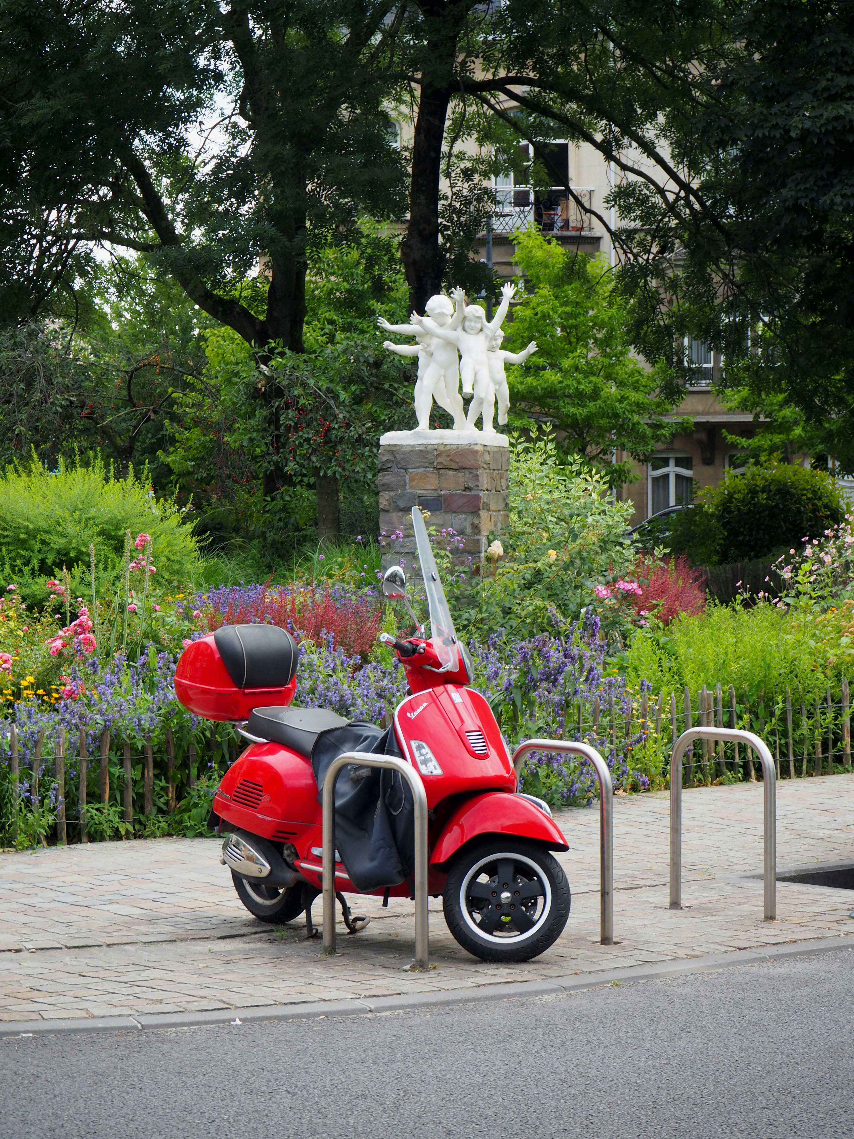 A vibrant red Vespa parked beside a colorful flower garden, with a whimsical sculpture of children playing in the background.
