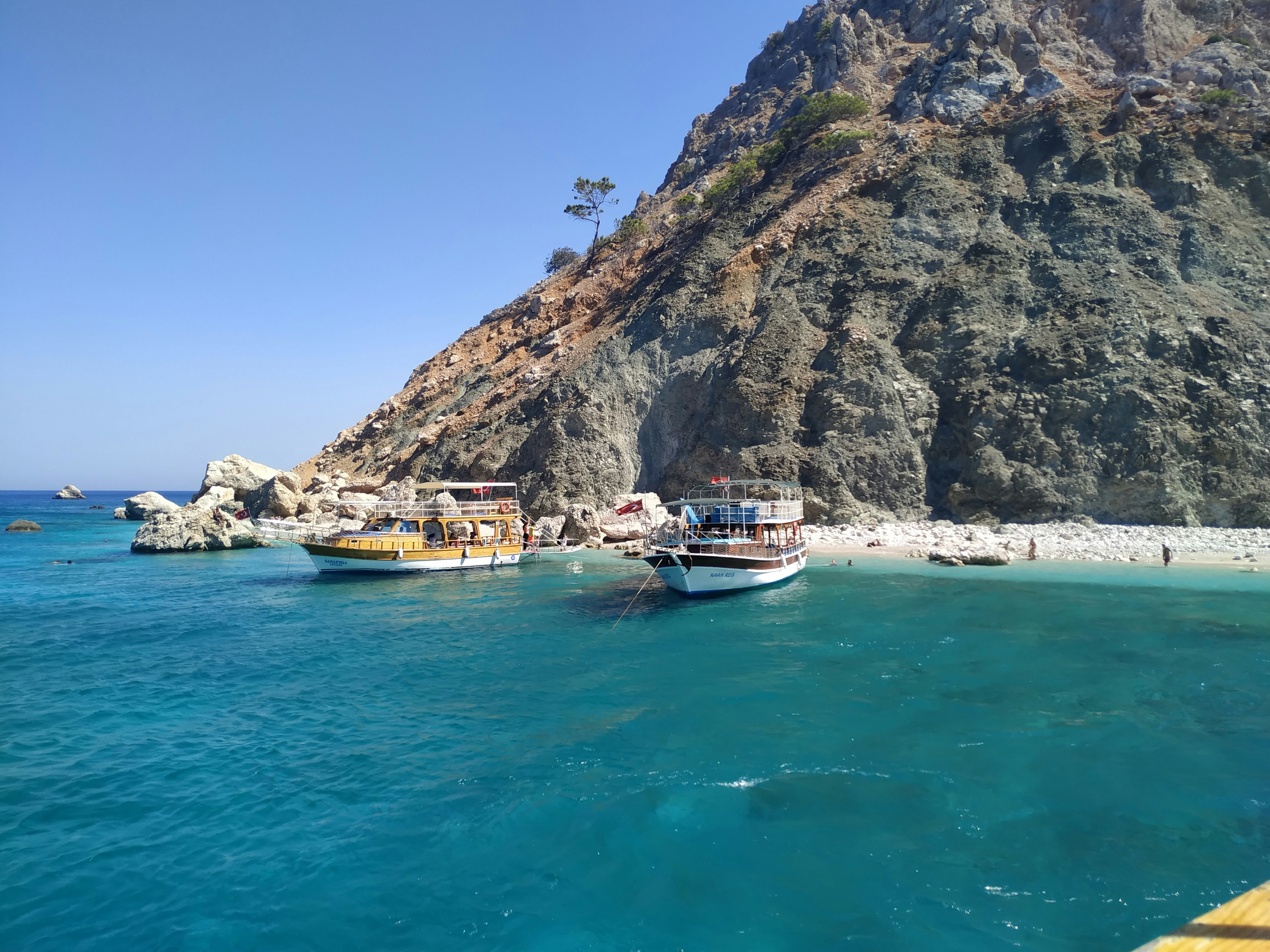 Boats anchored near a rocky cliff with clear turquoise water and a sandy beach under a bright blue sky.
