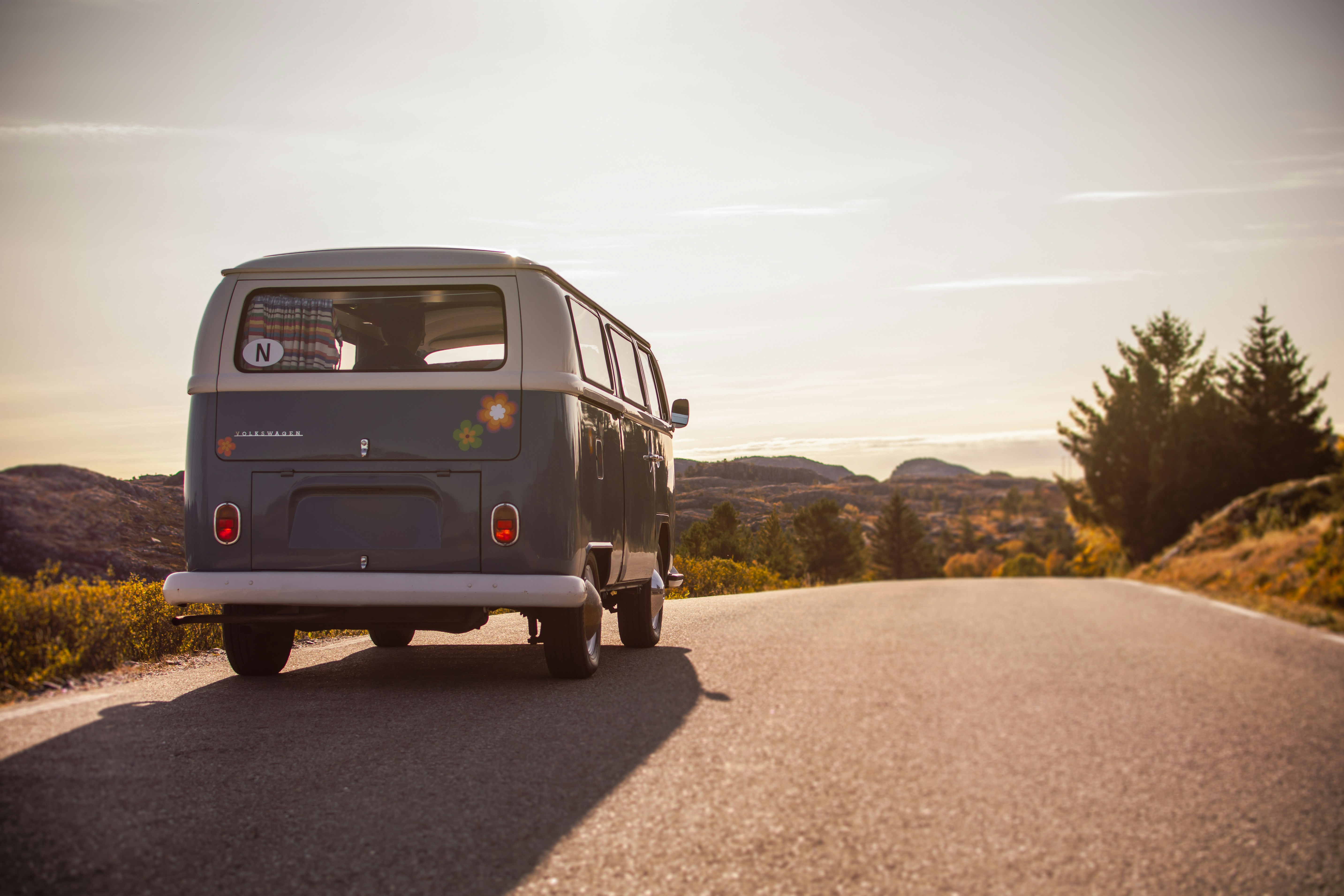 A classic blue Volkswagen van parked on a winding road, surrounded by rolling hills and trees under a soft sunset glow.