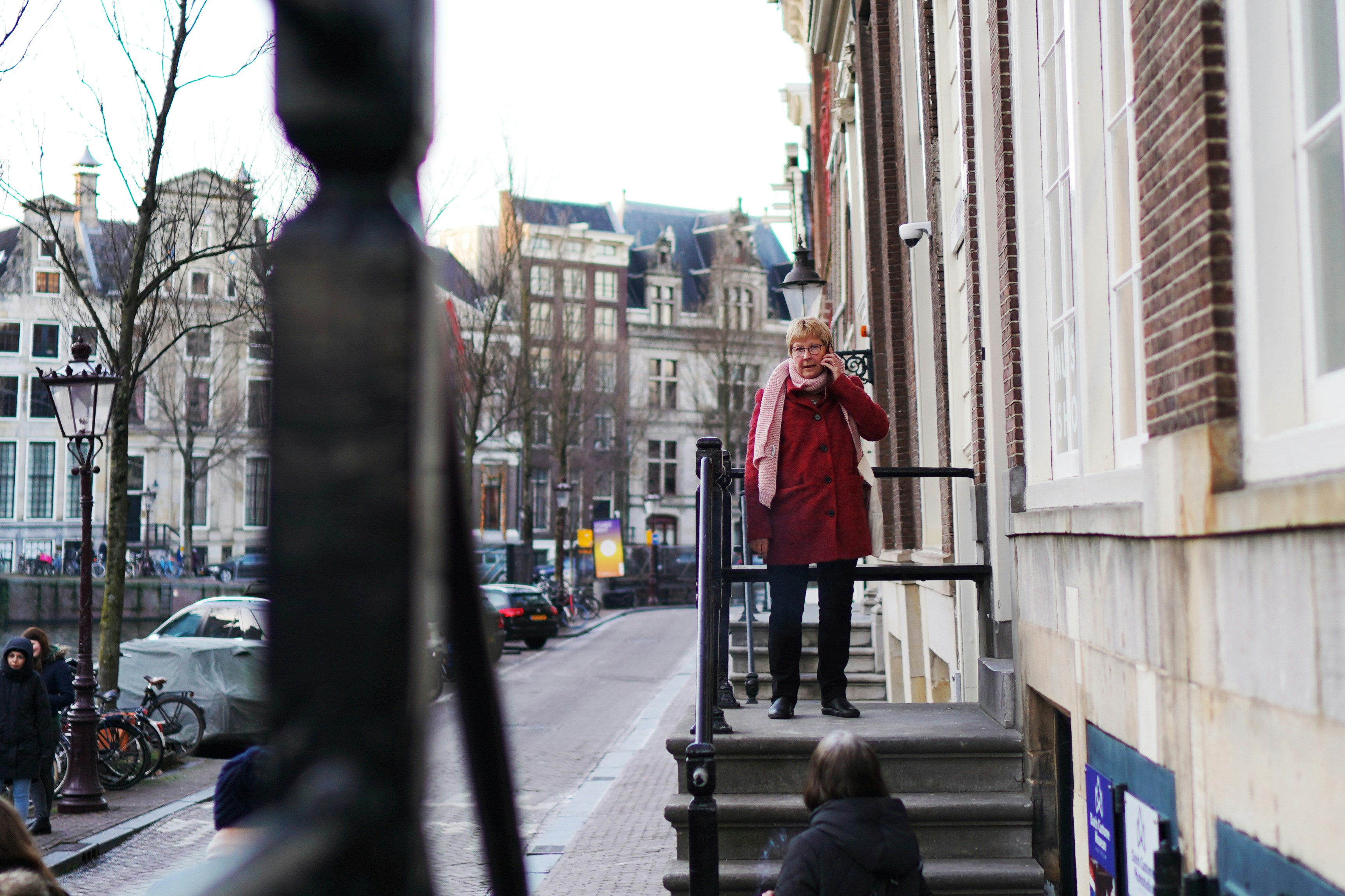 A woman in a red coat waves from a staircase in a bustling city street, surrounded by historic architecture and bicycles parked along the sidewalk.