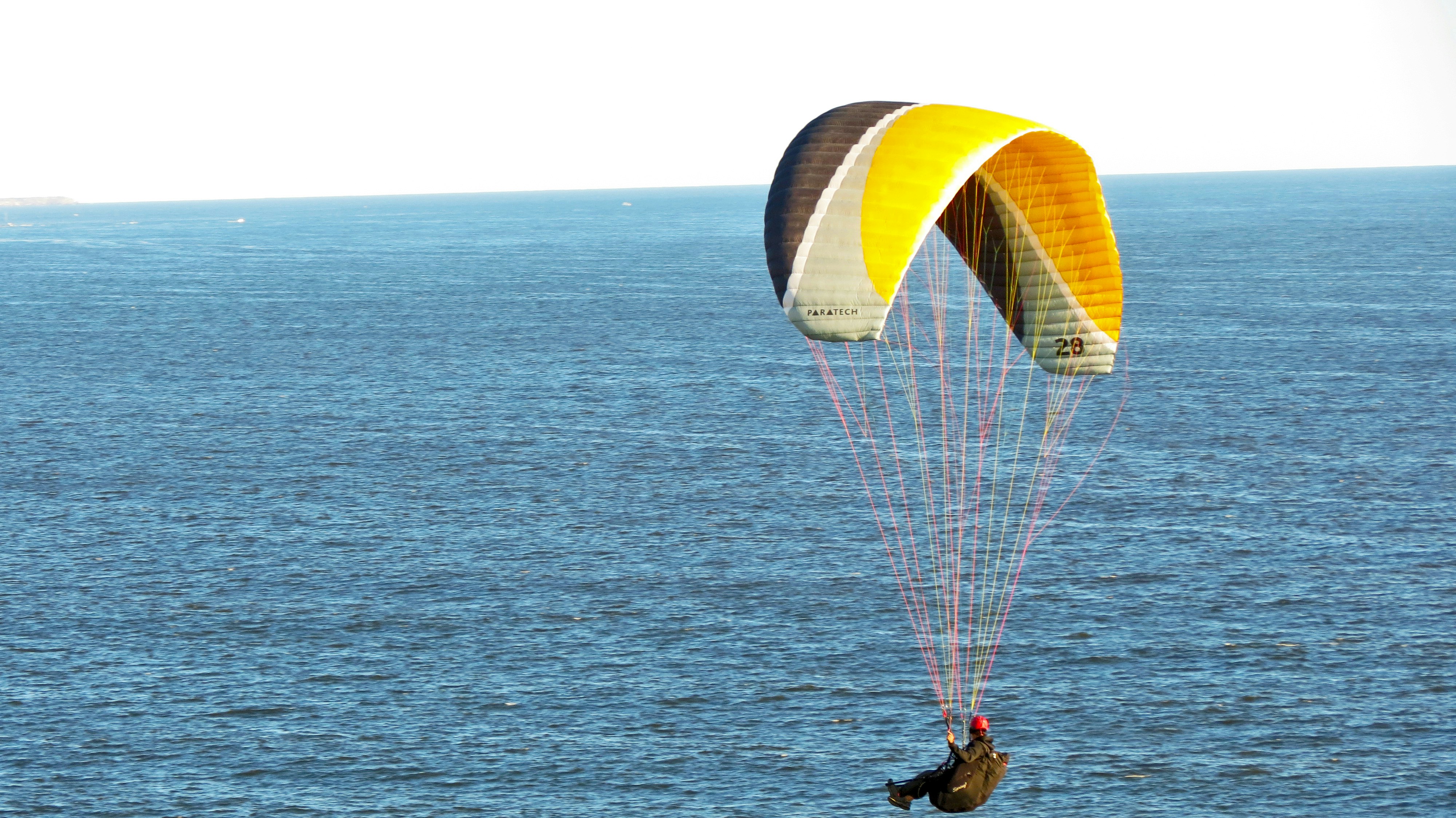 Paraglider in a black wetsuit soaring with a yellow and red parachute above the ocean during daylight.
