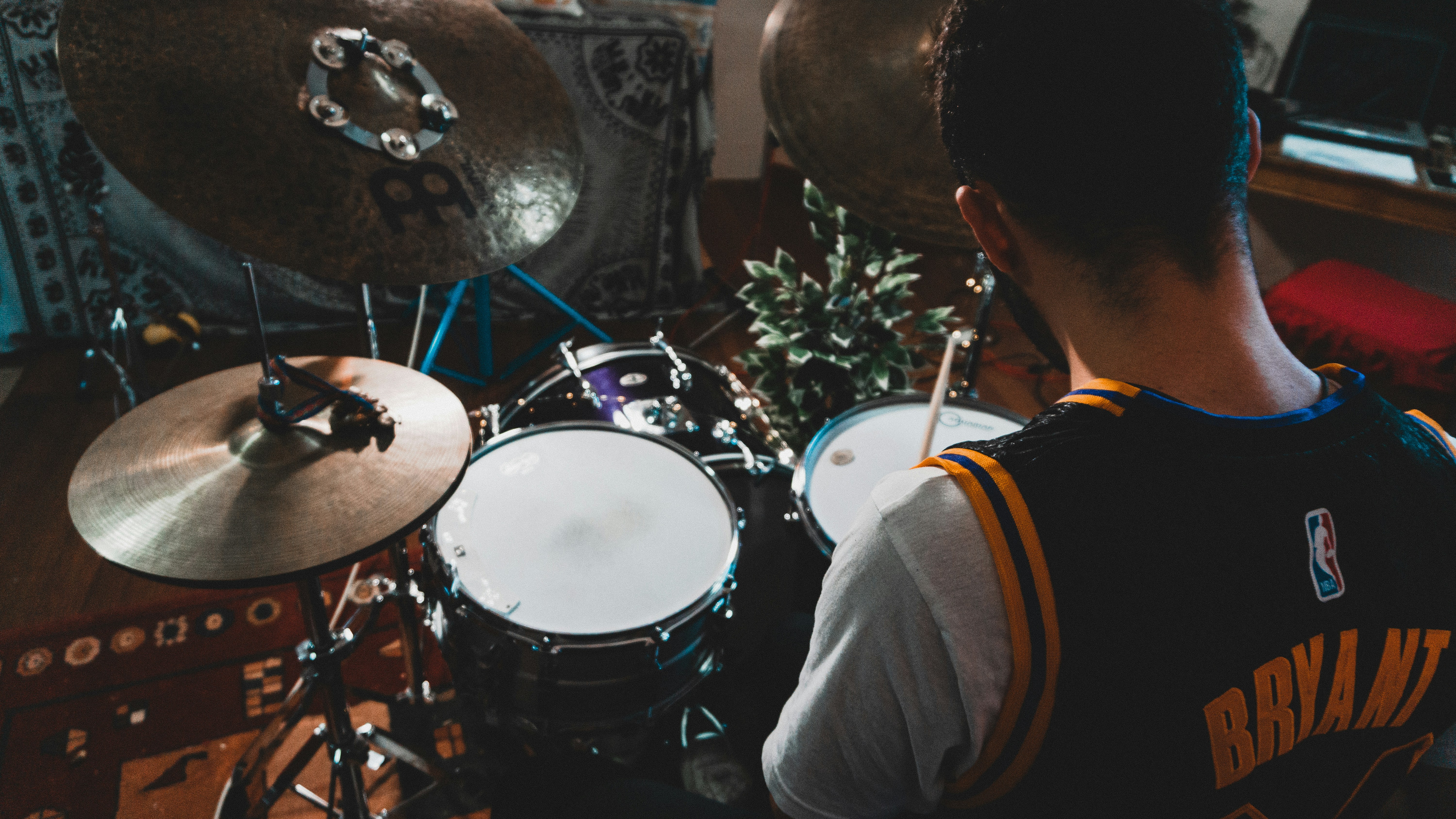 Drummer in a black jersey playing a drum kit, captured from behind with cymbals in view.