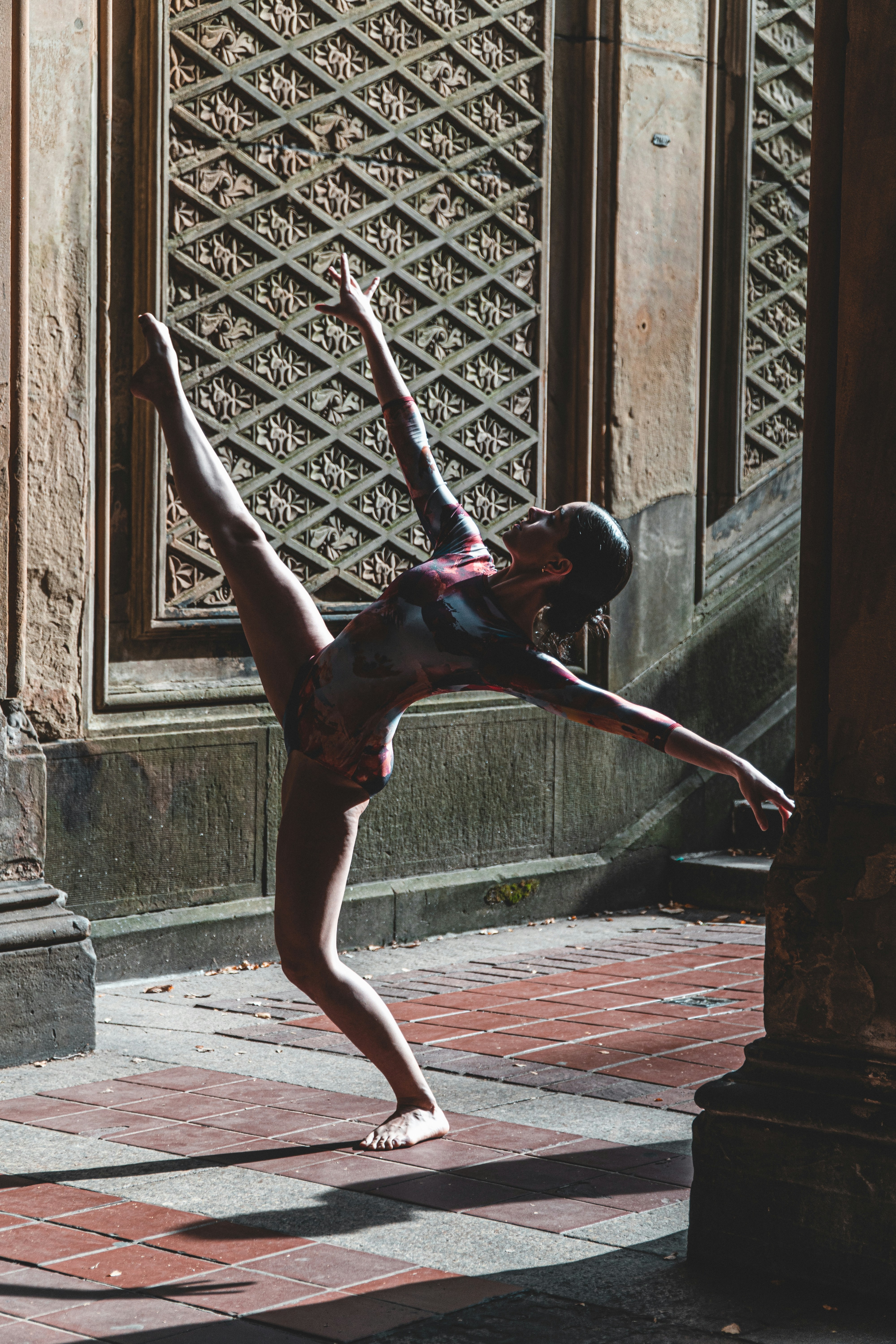 A dancer in a colorful bodysuit strikes a dynamic pose against an intricately patterned wall, illuminated by soft natural light. The contrasting shadows enhance the fluidity of the movement.