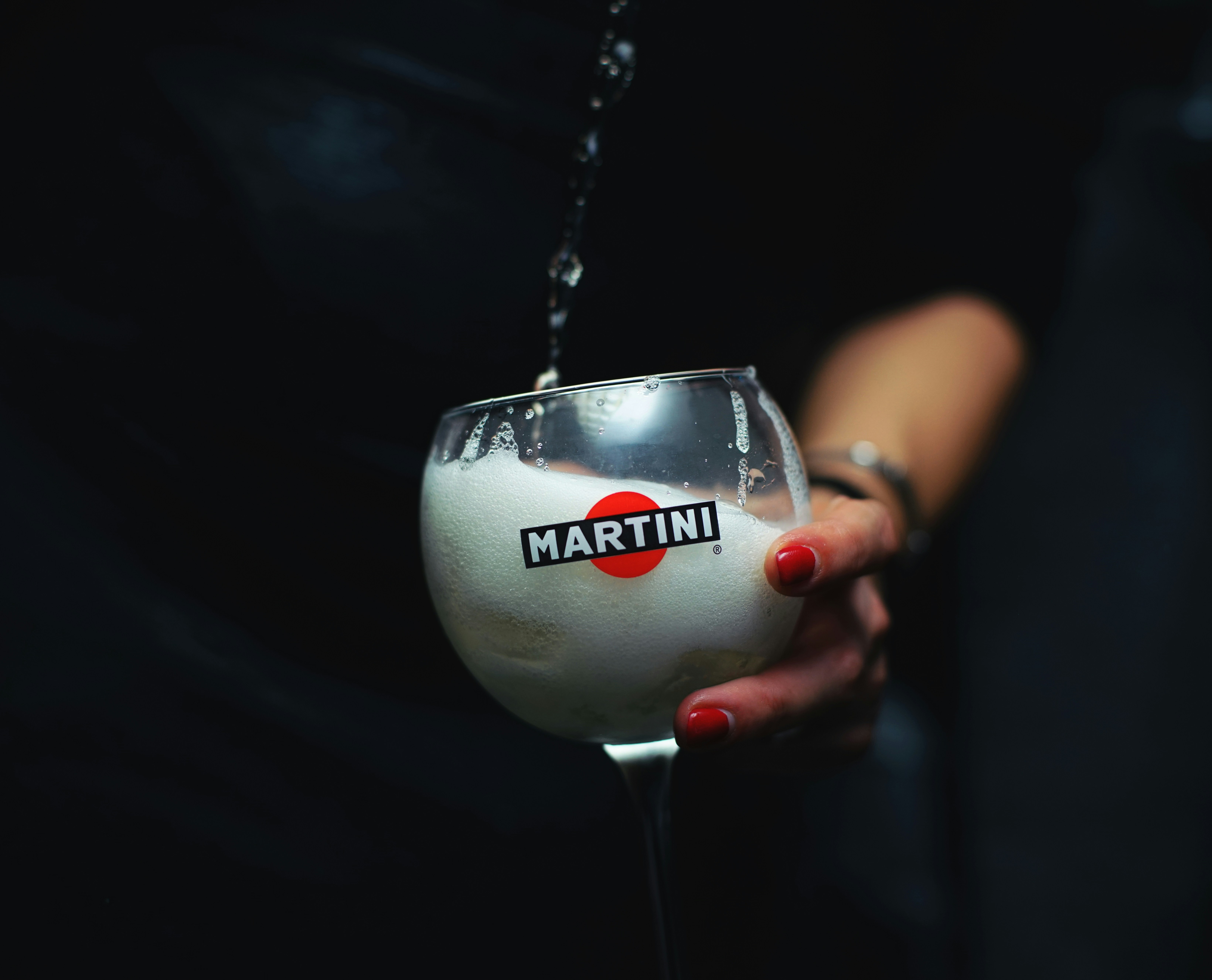 A hand pours a sparkling drink into a MARTINI-branded glass, showcasing the frothy liquid and the logo prominently. The background is softly blurred, emphasizing the drink's detail.