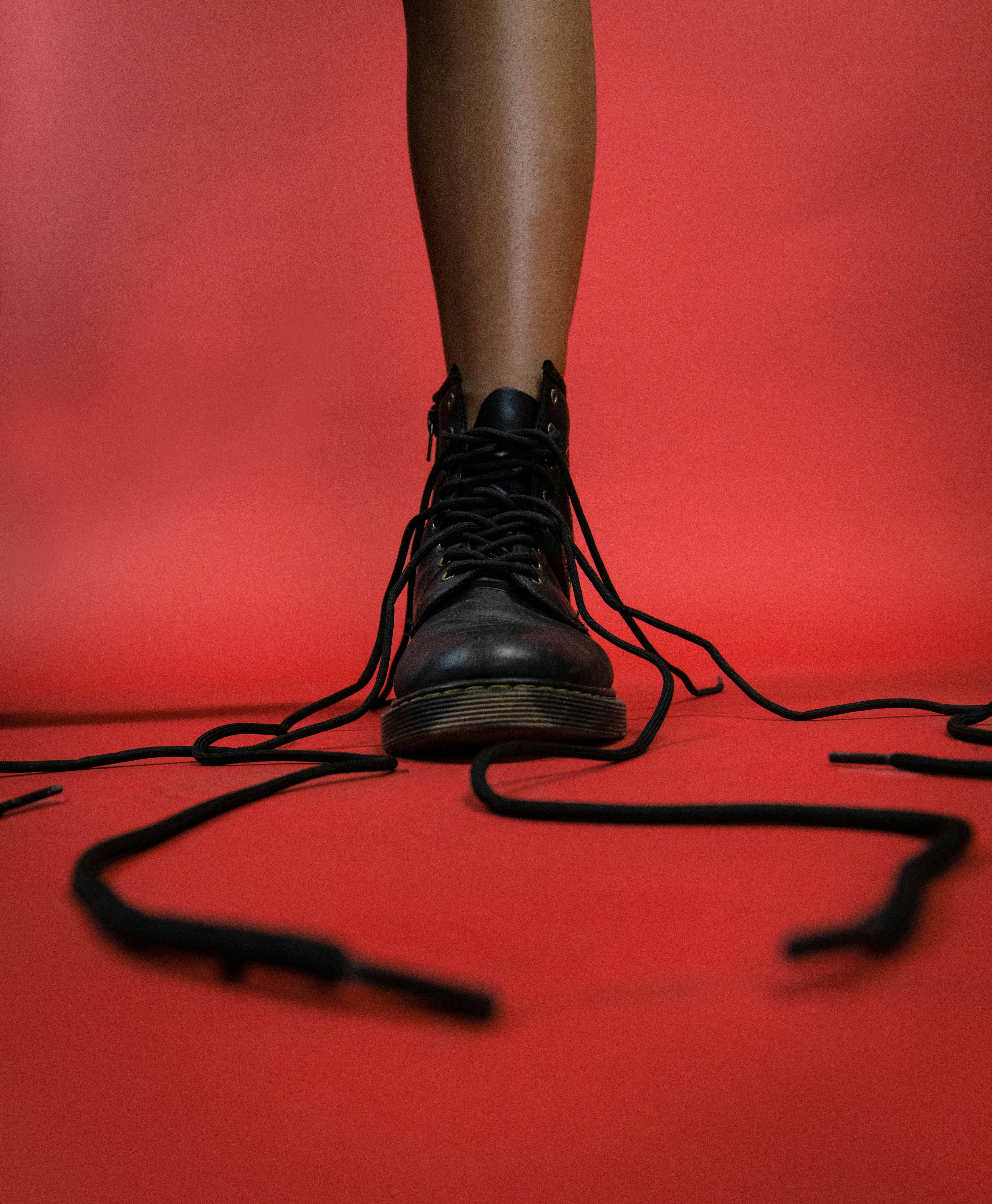 Close-up photograph of a black combat boot stepping toward the camera on a red backdrop, with tangled laces sprawling across the floor.