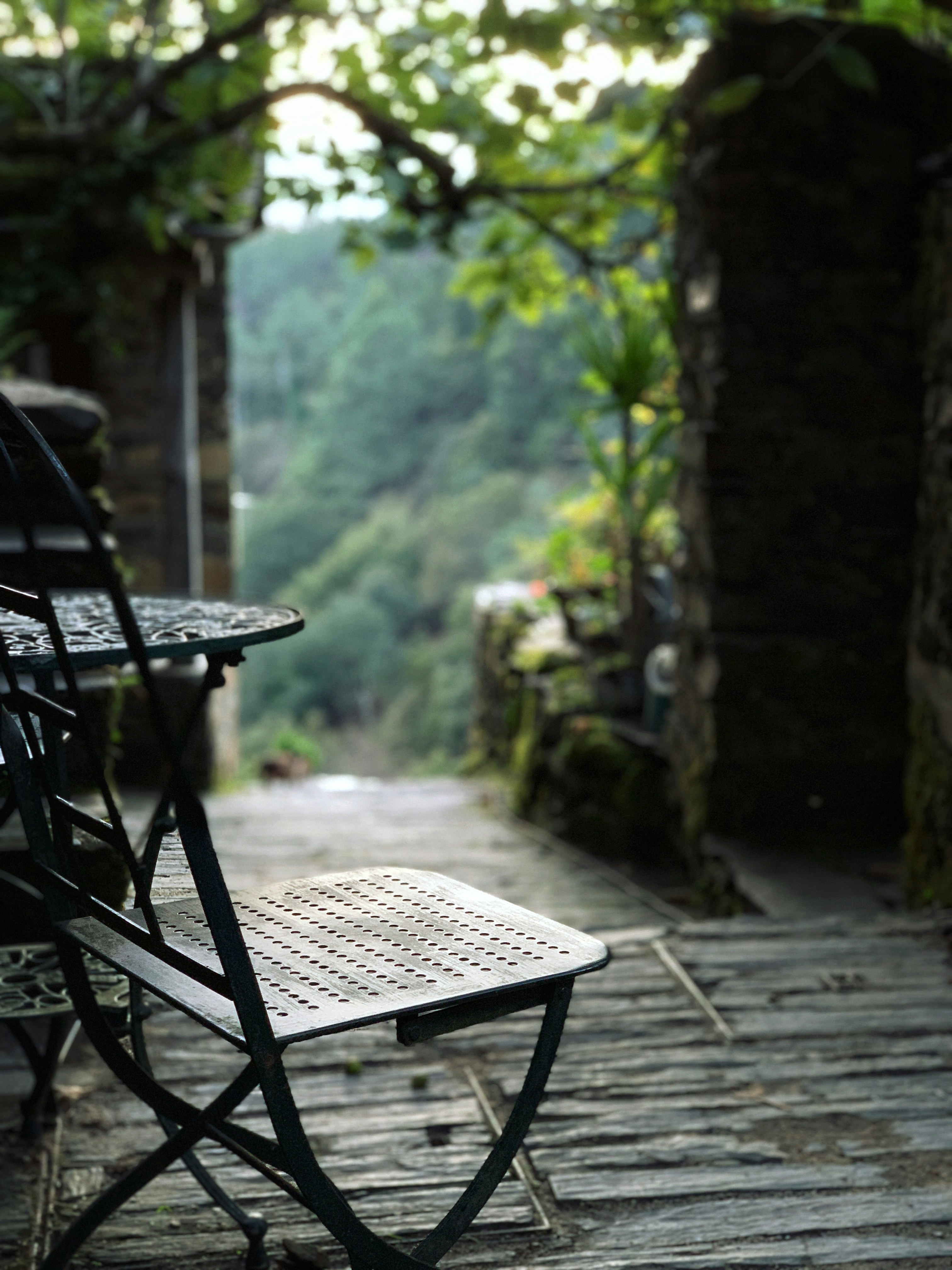 An intricately designed iron chair sits on a rustic stone pathway, framed by lush greenery and a distant view of rolling hills. The scene evokes a sense of calm and contemplation.