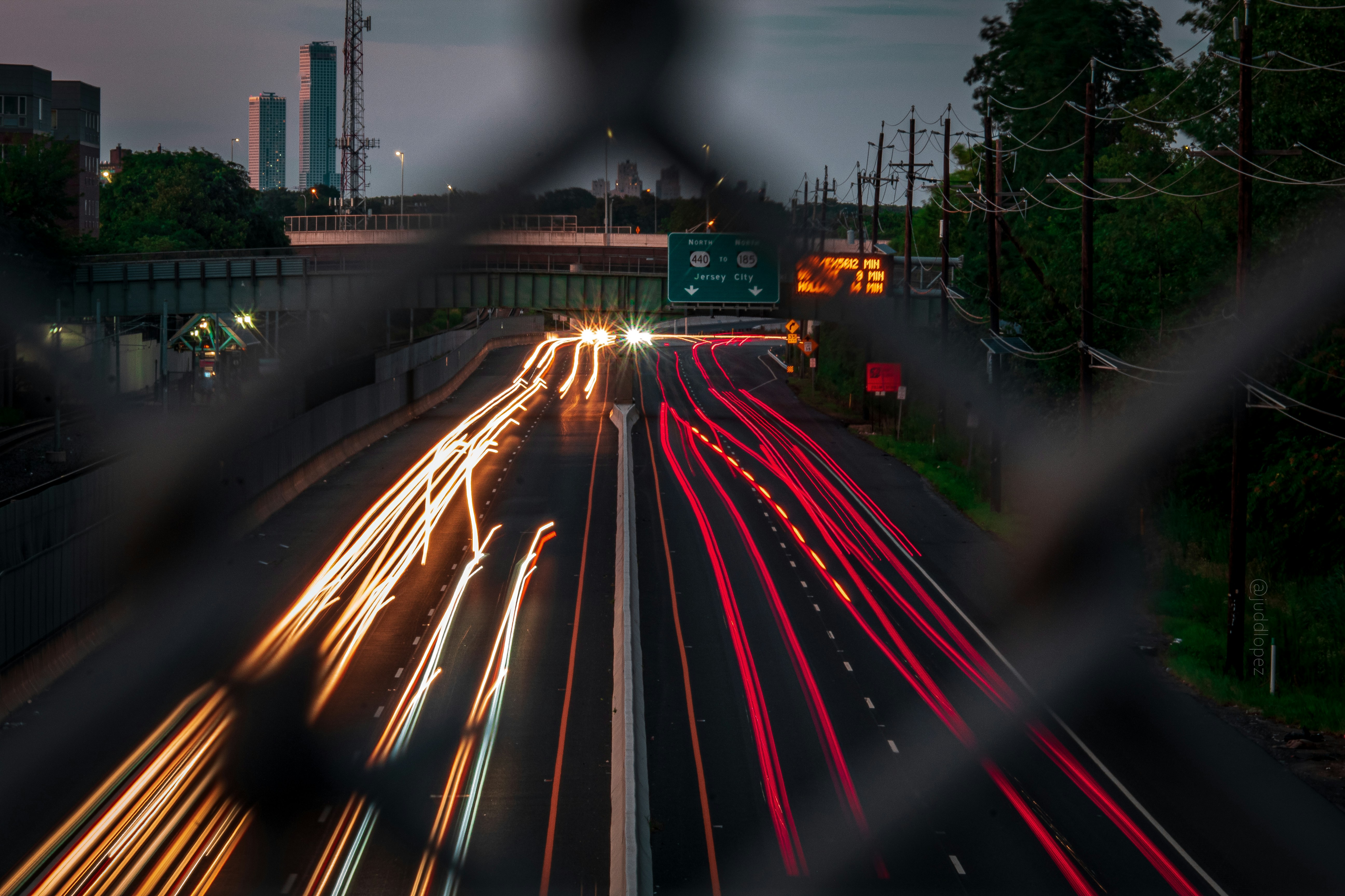 Long exposure of car light trails on a highway viewed through a chain-link fence at dusk.