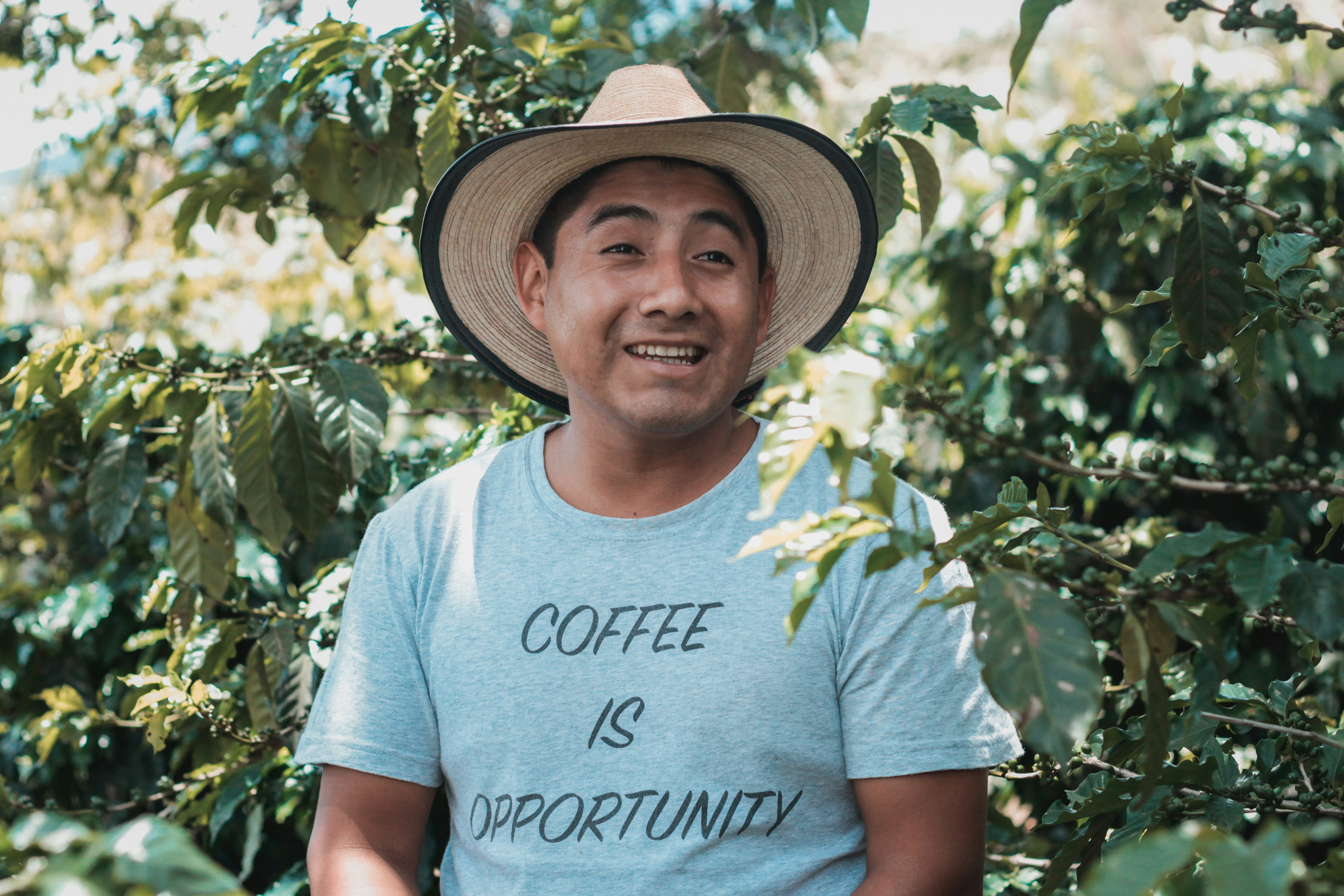 Smiling man in a hat stands among lush coffee plants wearing a shirt reading 'Coffee is Opportunity.'