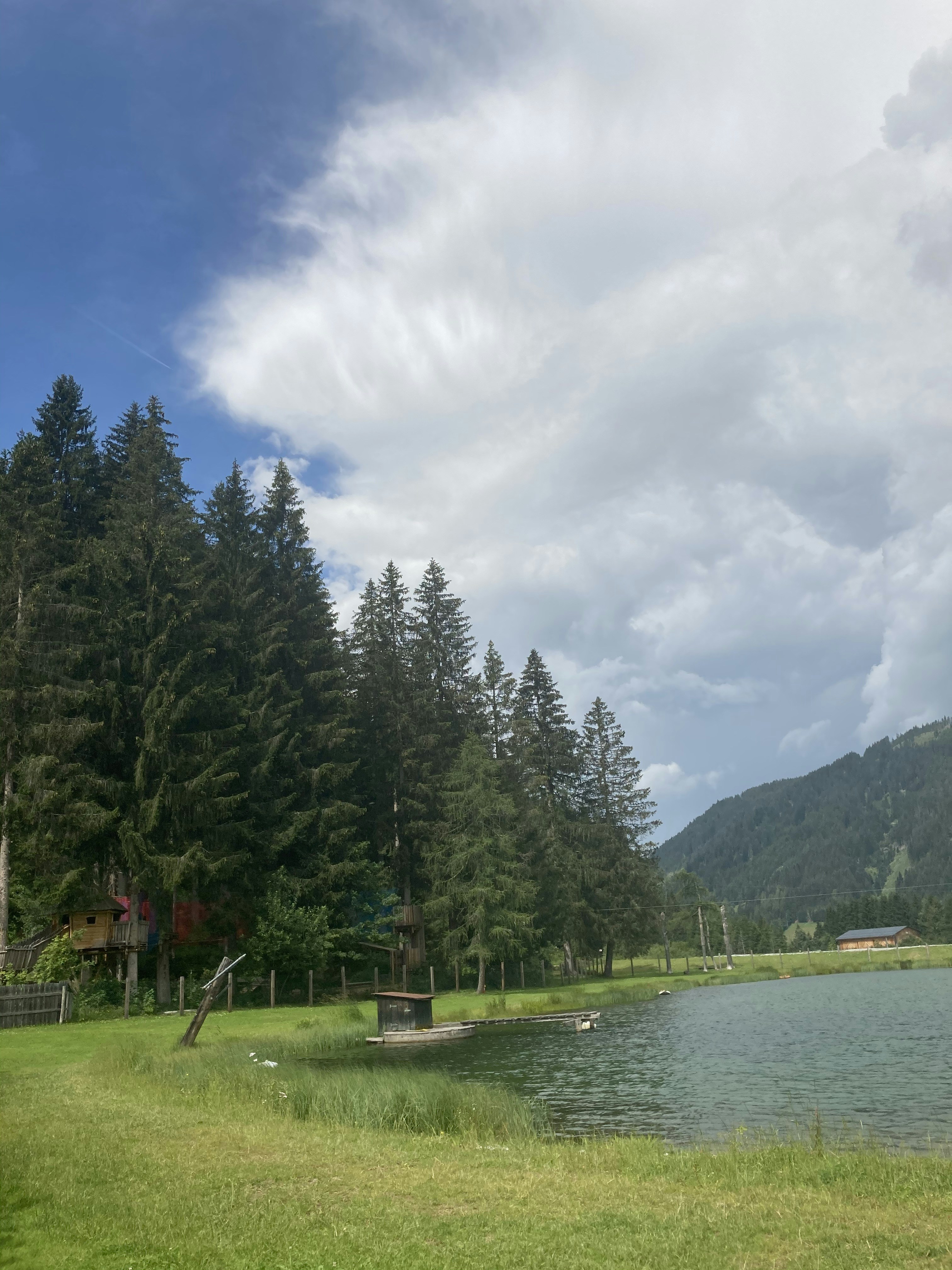 Lush green trees lining a tranquil lake under a dynamic sky filled with clouds. A small dock extends into the water, inviting reflection.