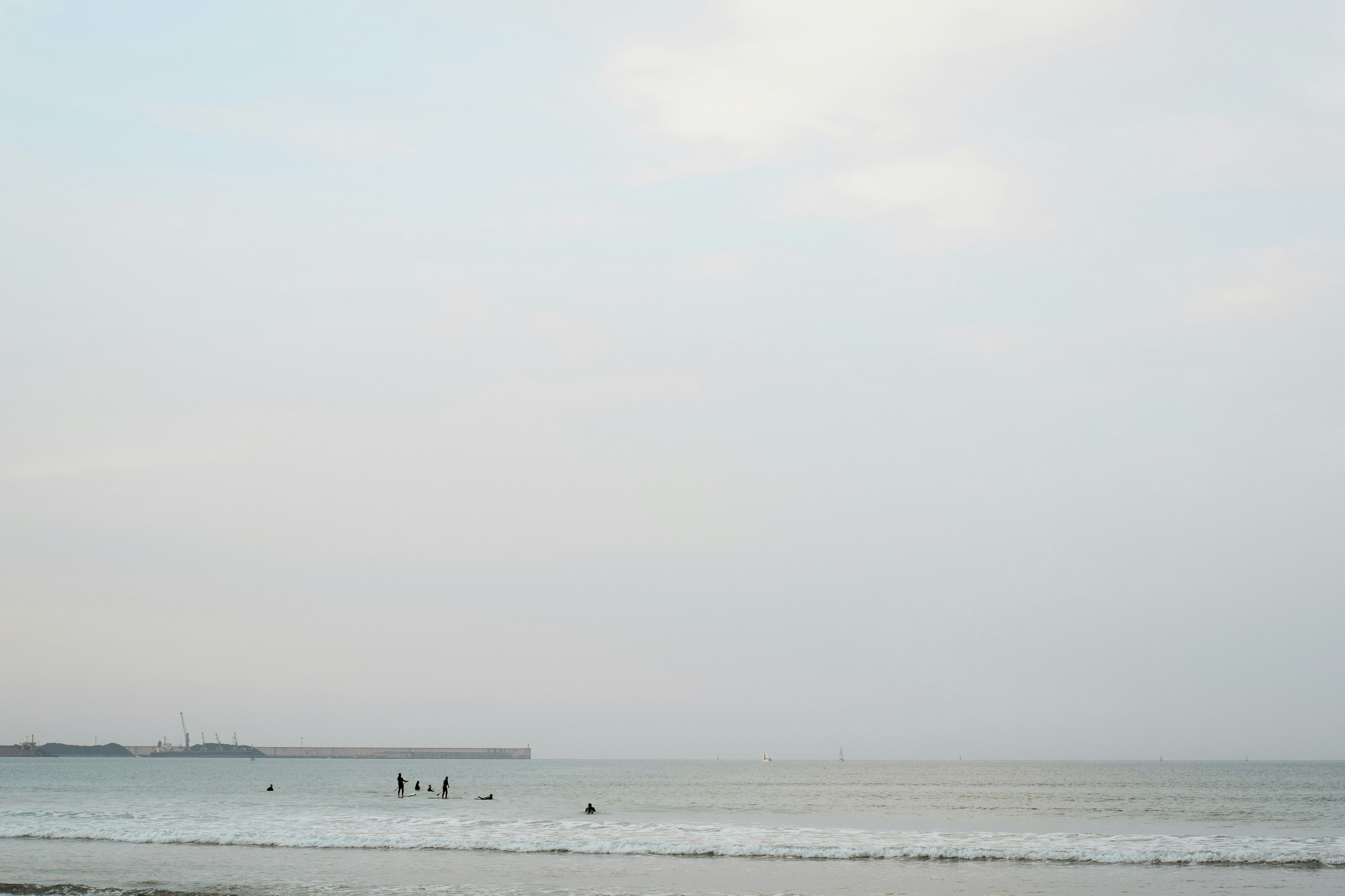 Surfers enjoying gentle waves under a pastel sky, with a distant shoreline hinting at human presence. 