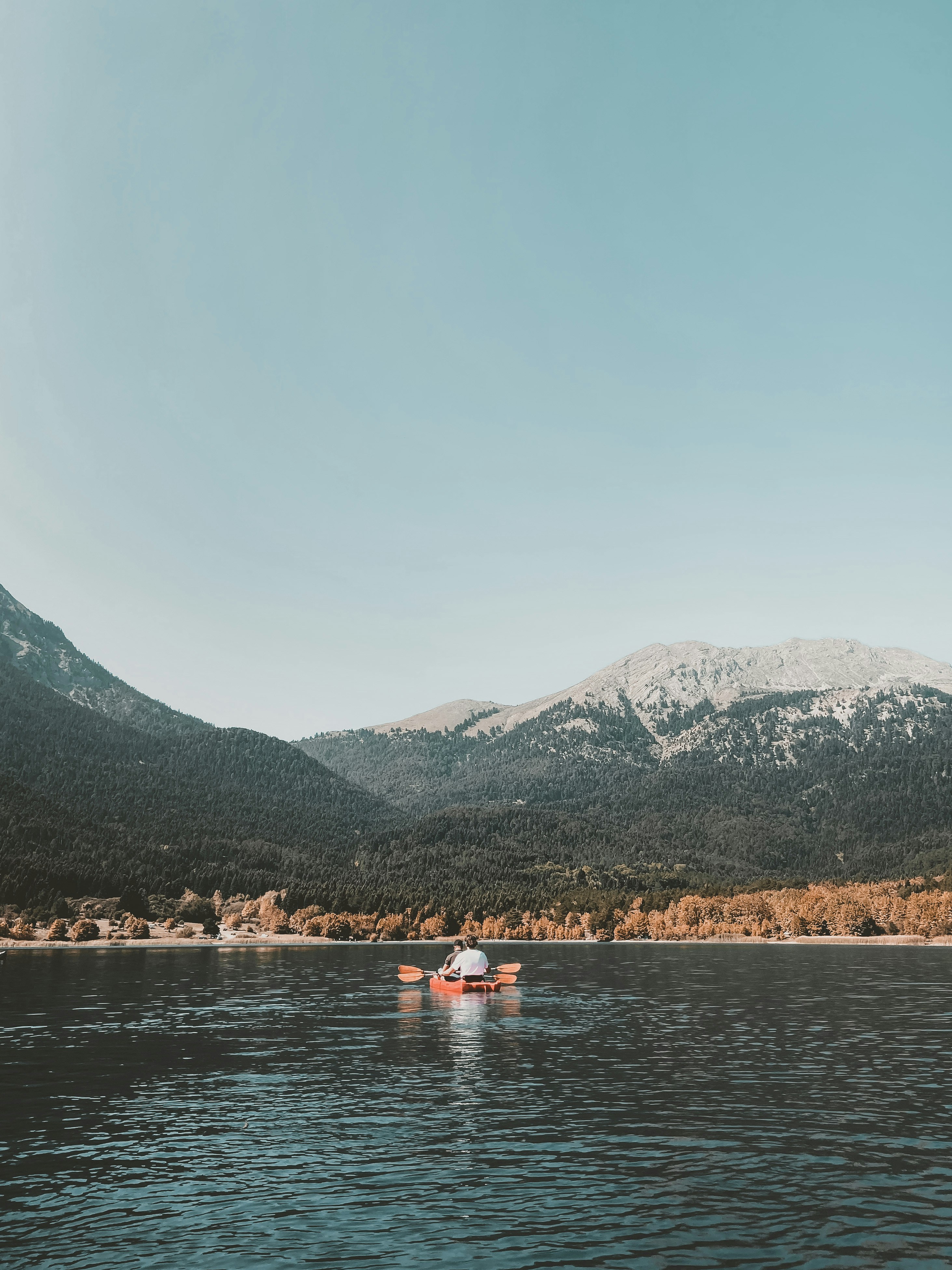 A lone kayaker glides across a tranquil lake surrounded by majestic mountains and lush forests. The clear blue sky reflects off the water's surface.