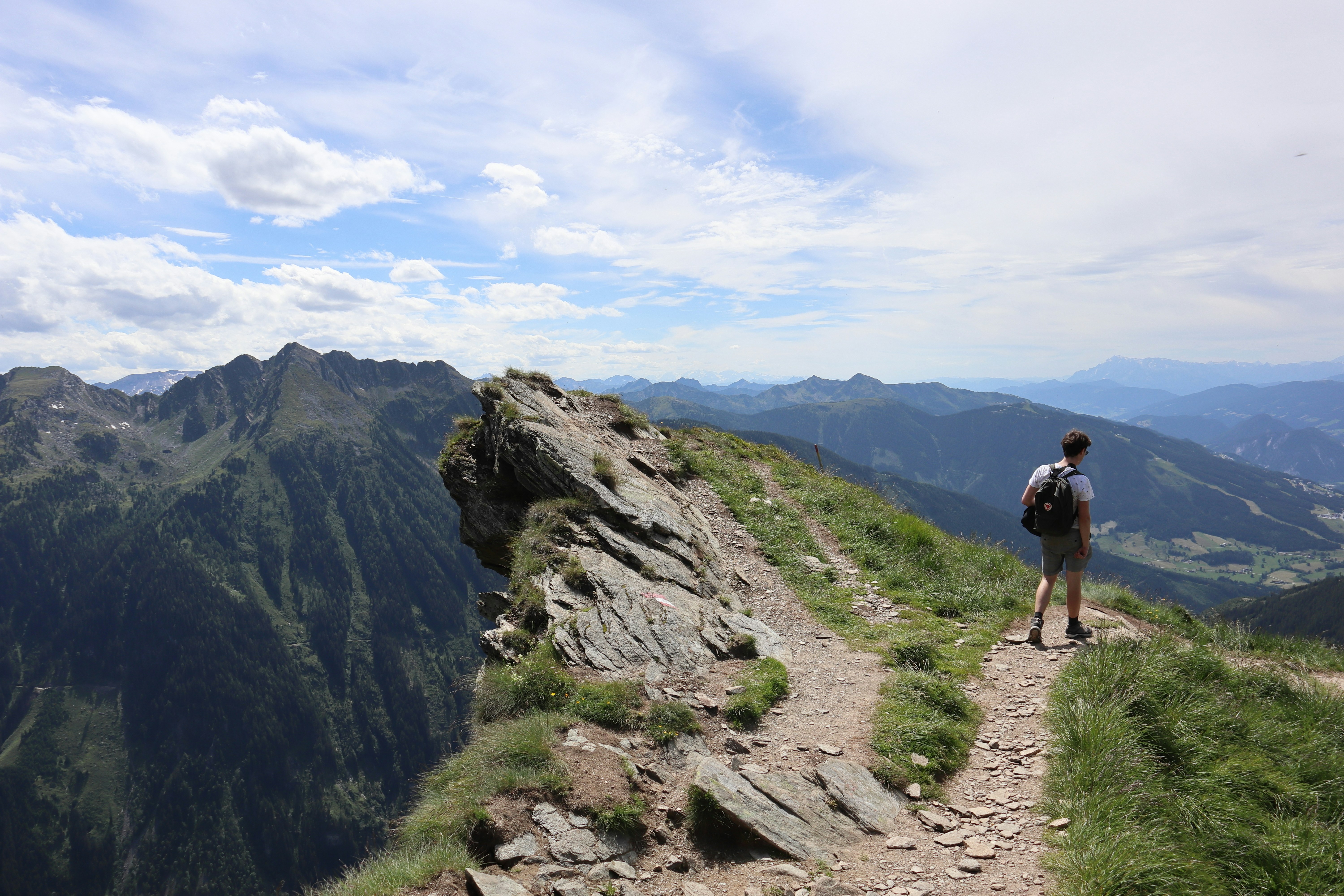 Hiker standing on a rocky ridge overlooking expansive mountain ranges under a partly cloudy sky.
