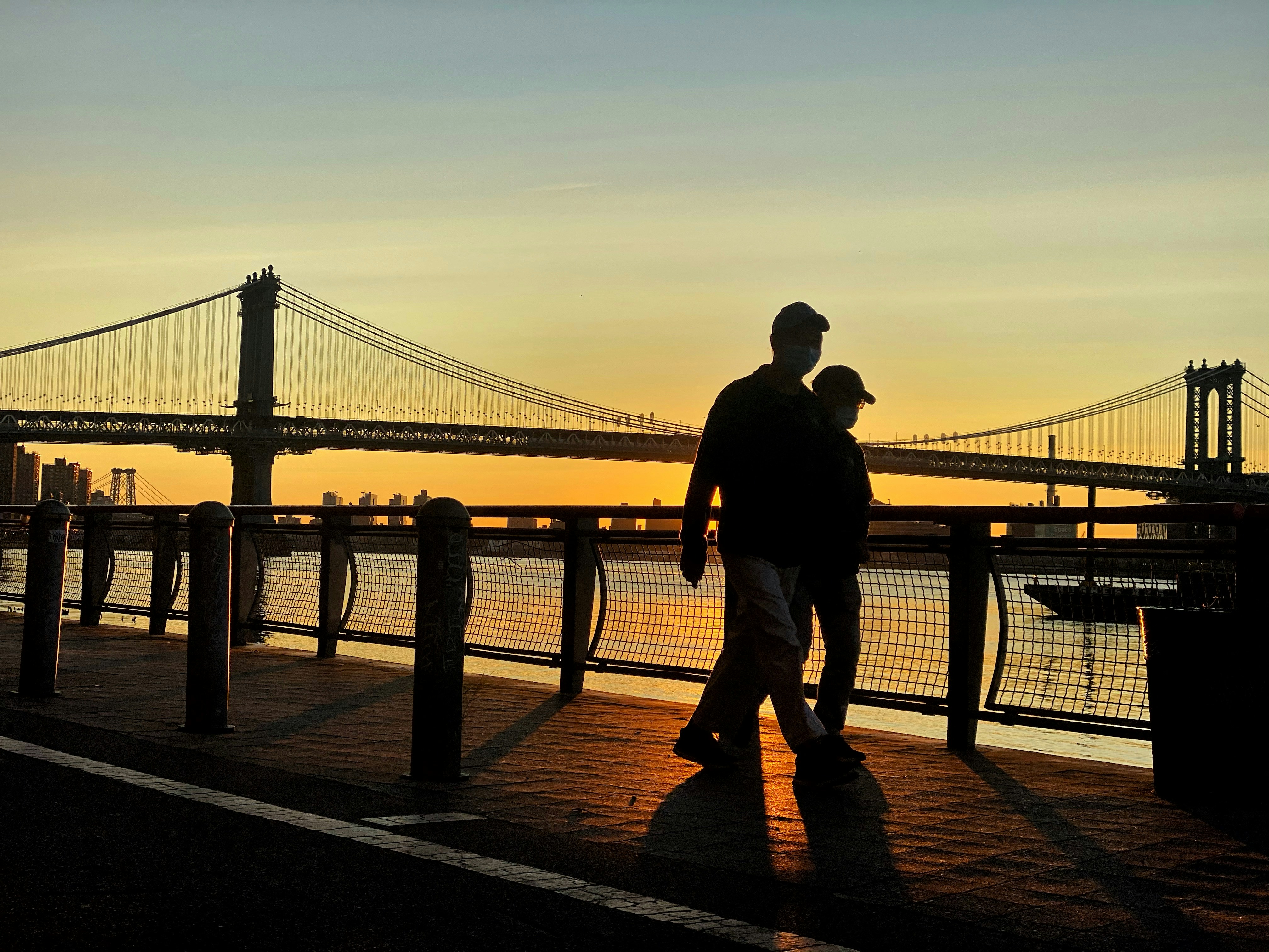 Silhouetted figures walking along a waterfront with a bridge and sunrise in the background.