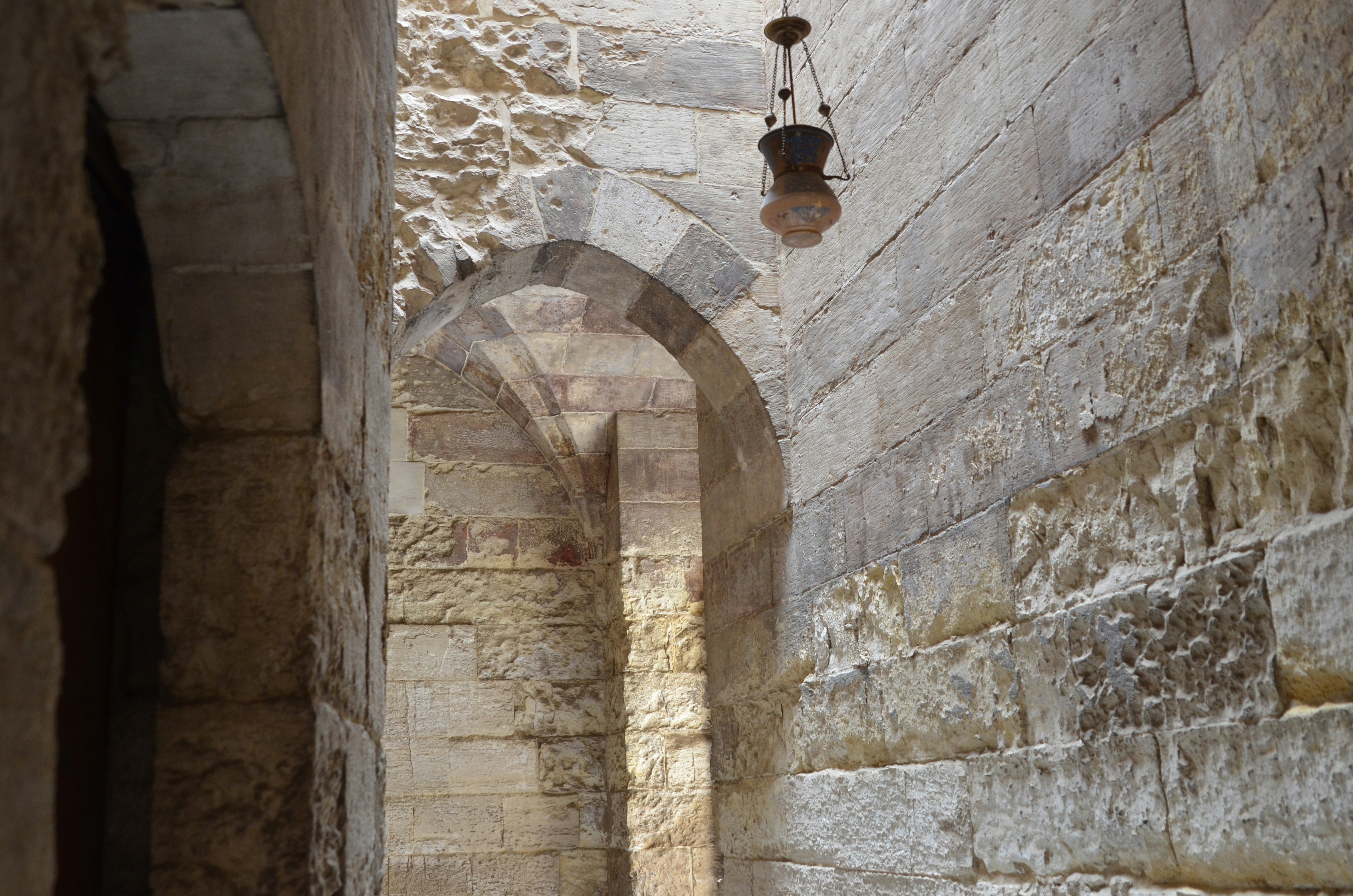 Ancient stone corridor illuminated by a hanging lantern, showcasing textured walls and archways. The scene evokes a sense of timelessness.