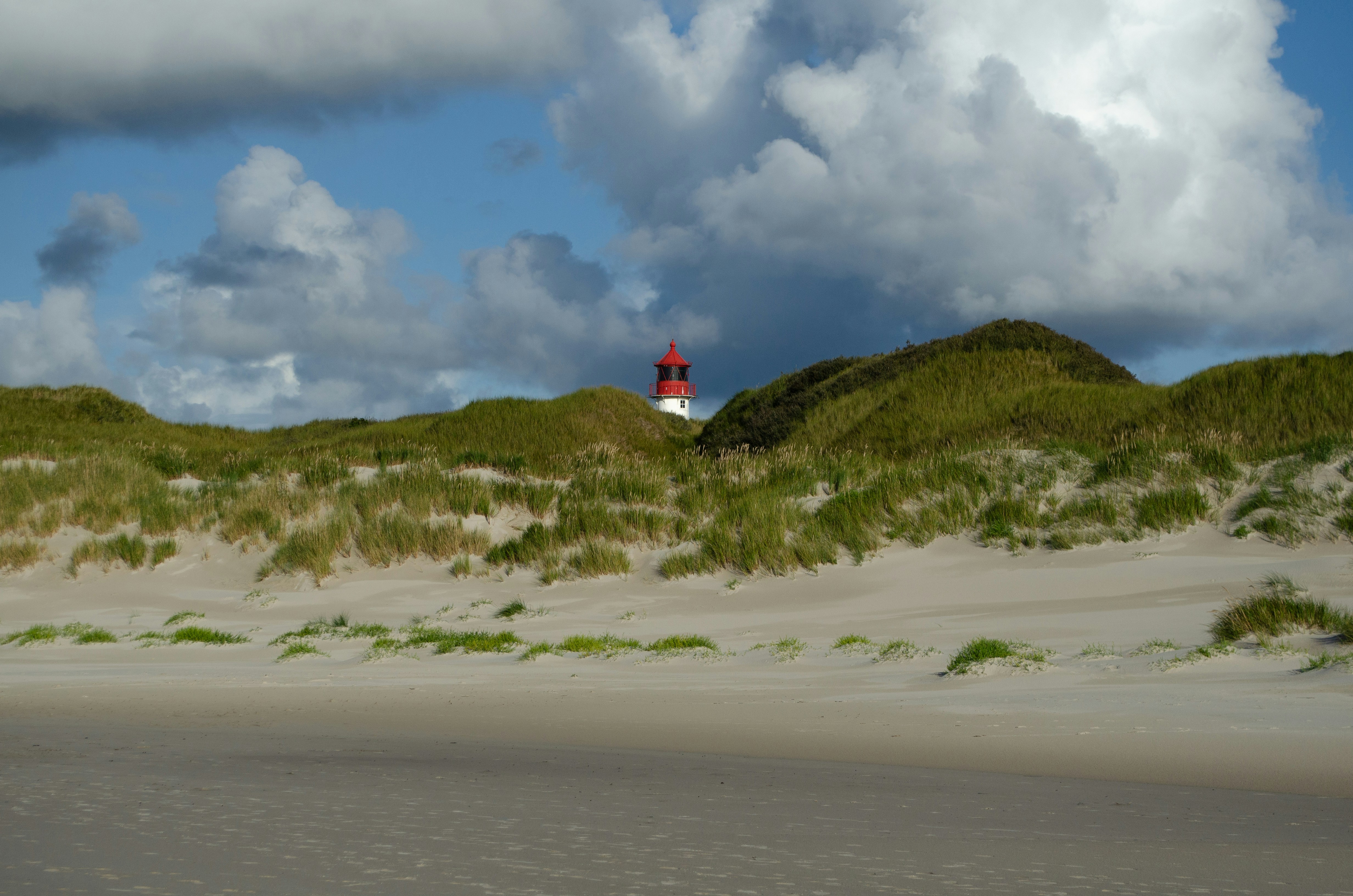 Lighthouse peeking through grassy dunes under a cloudy sky, highlighting the serene coastal landscape.