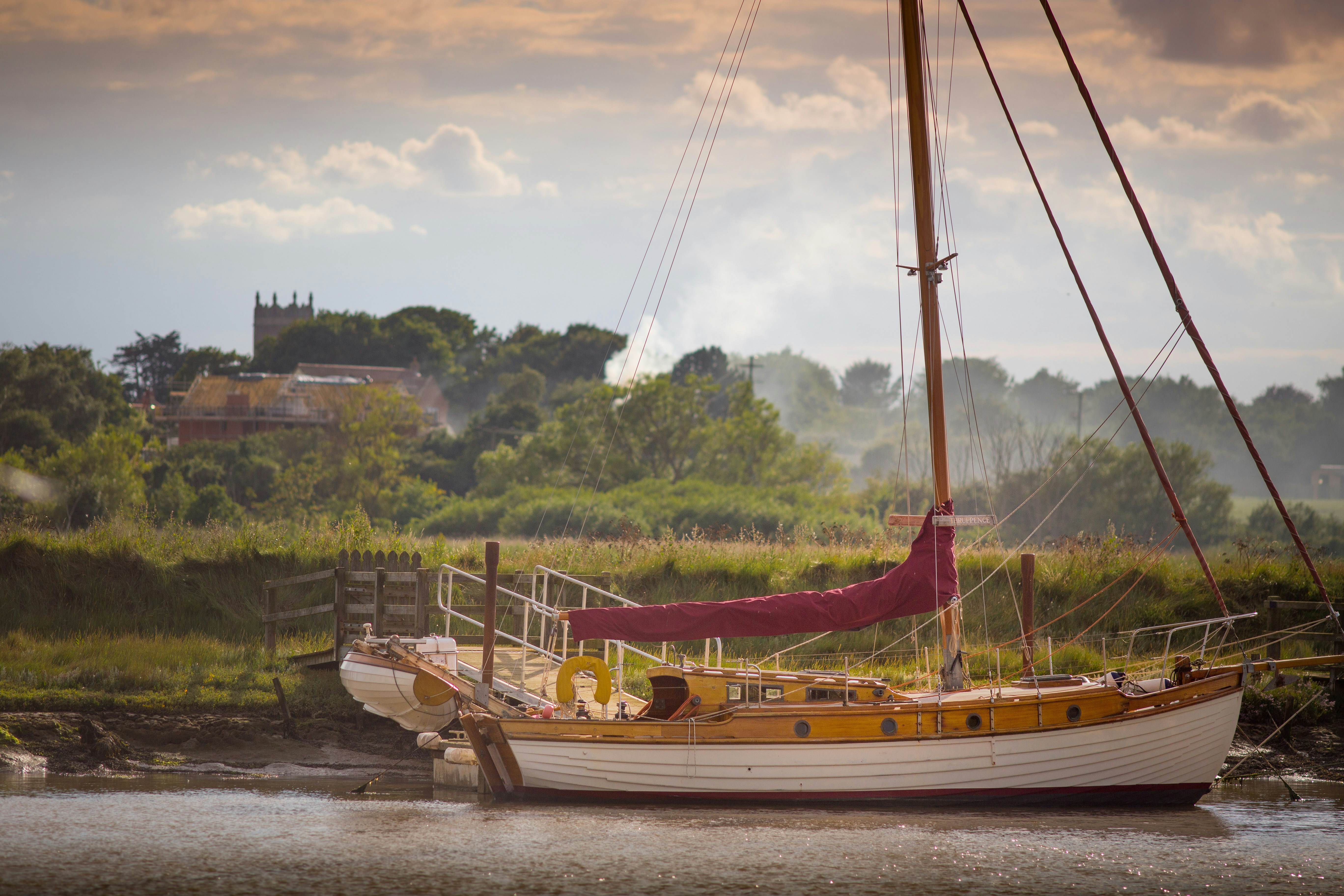 Wooden sailboat moored by a lush riverside with a distant church tower under a cloudy sky.