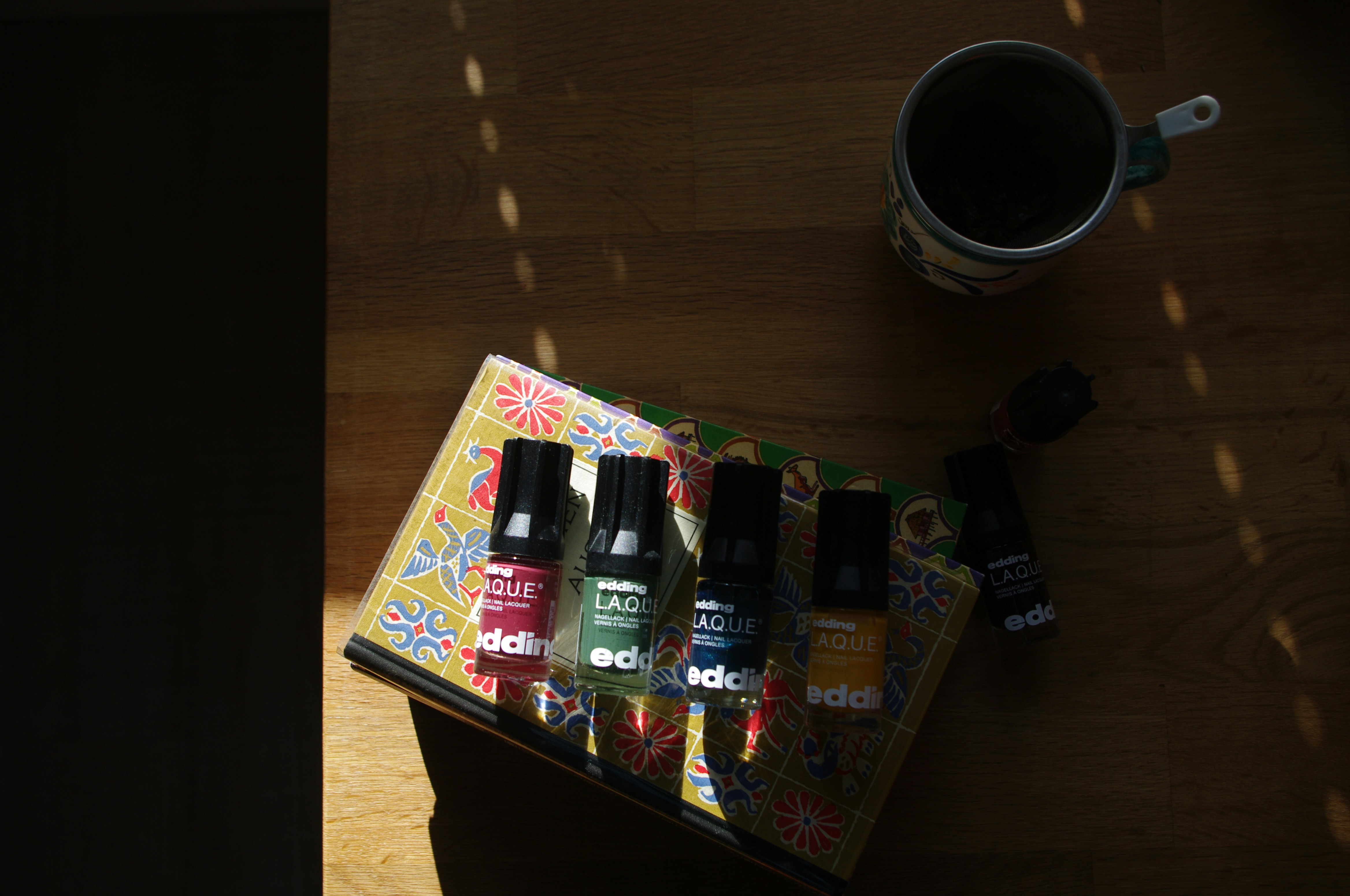 Five small color bottles sit on a yellow floral tin on a sunlit wooden table, with a mug in the background.