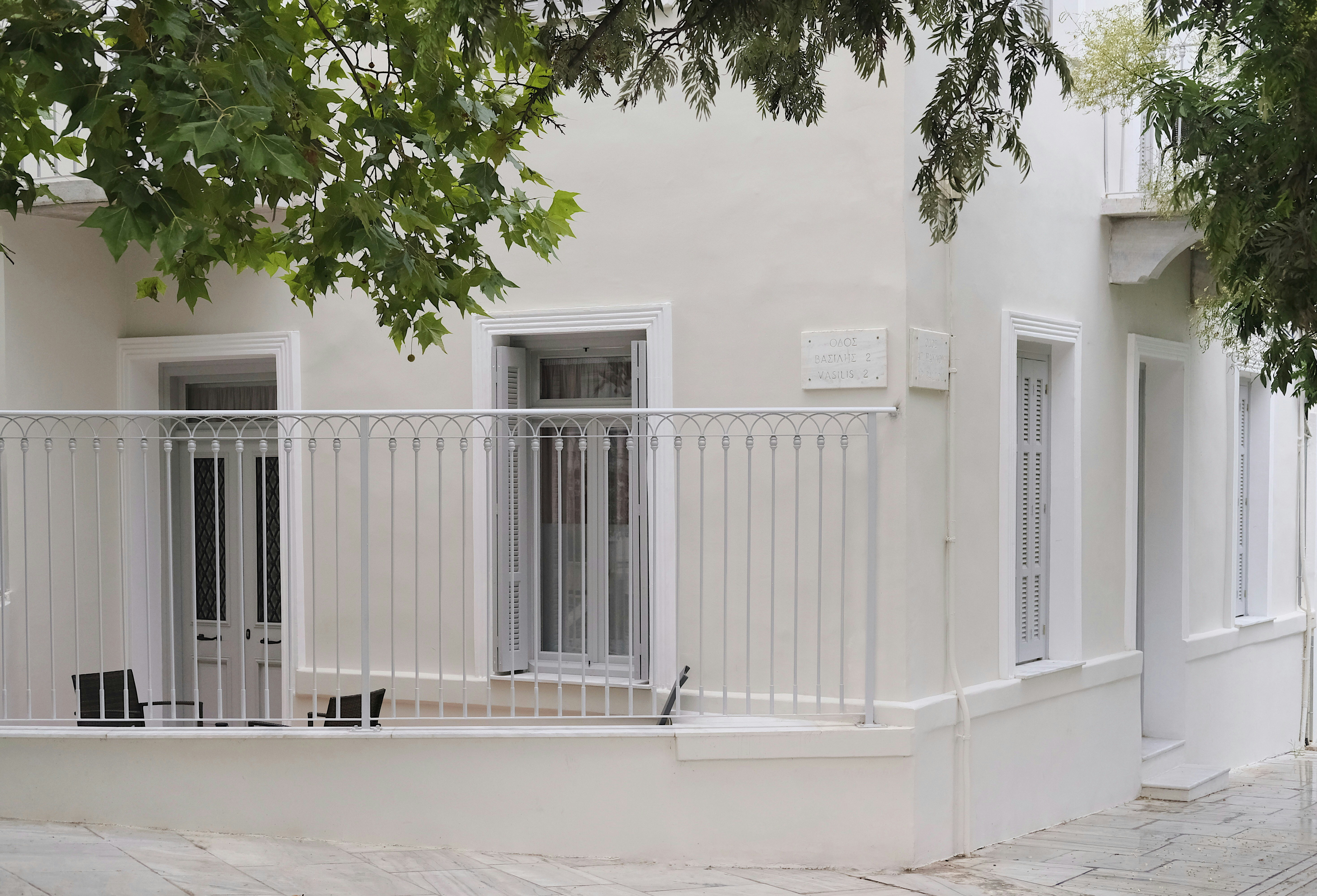 Charming white building with elegant iron railing and large windows, framed by lush greenery. The simplicity of the design invites contemplation.