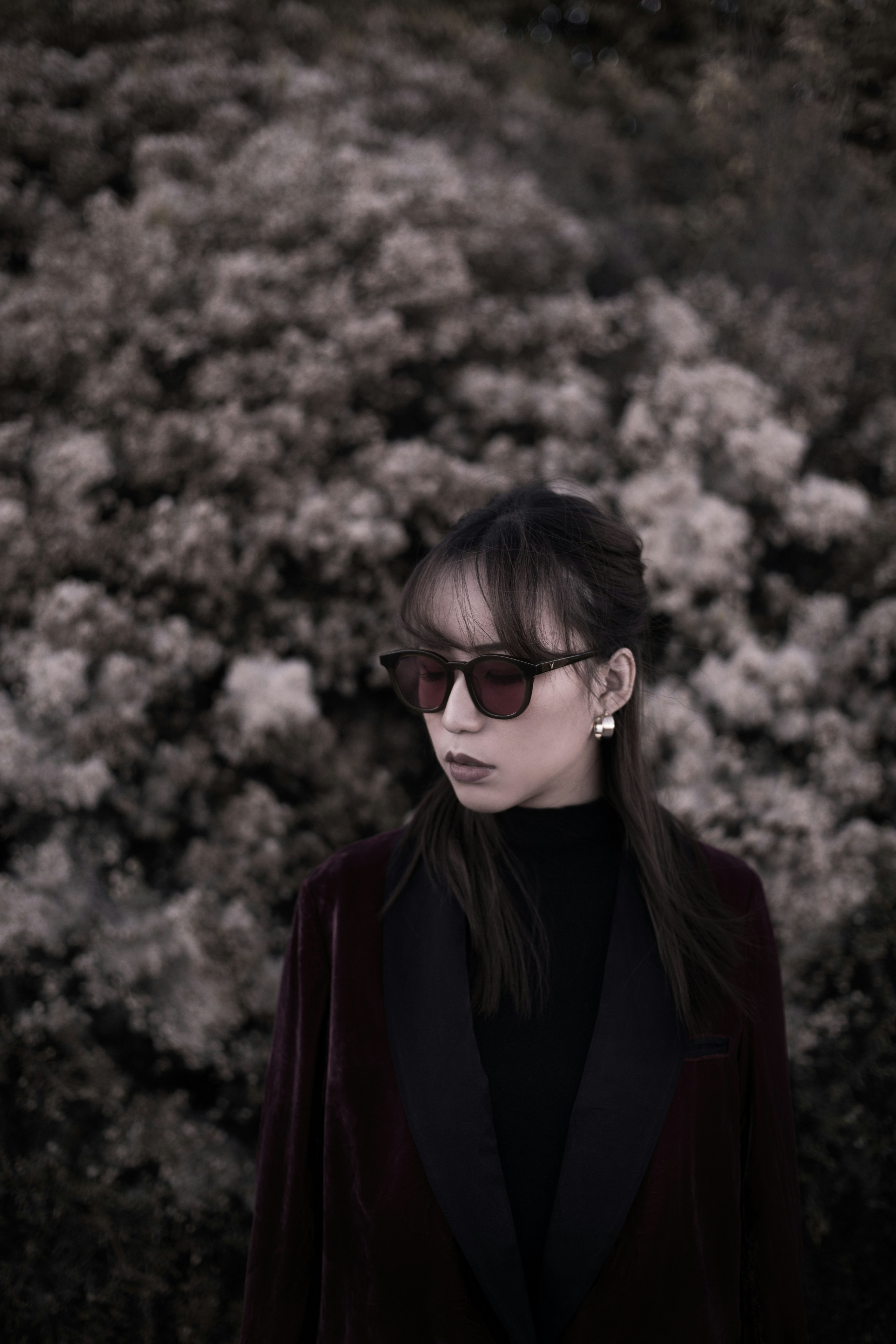 Woman in sunglasses stands against a backdrop of dense, muted foliage.