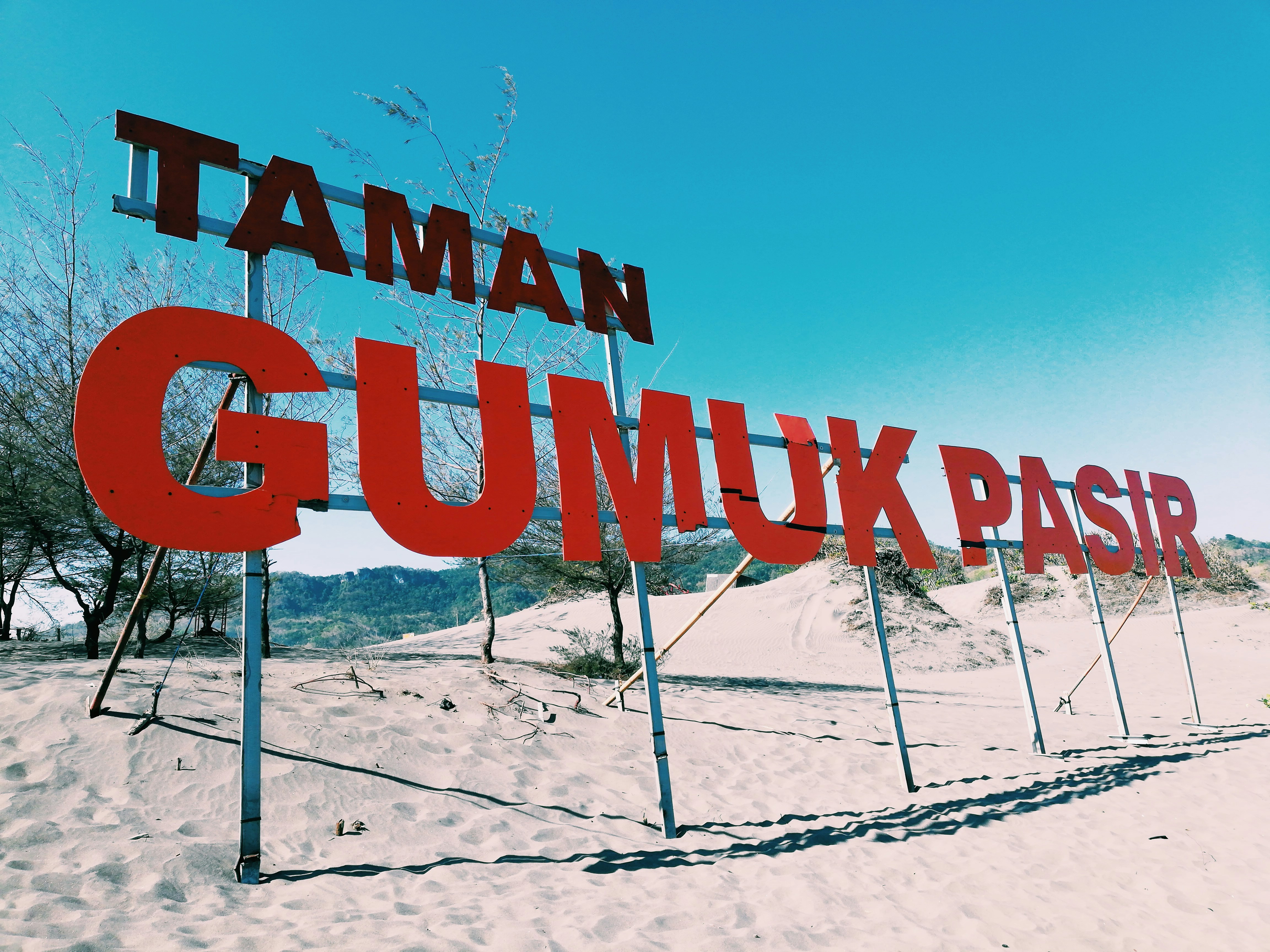 Large red letters spell out 'Taman Gumuk Pasir' against a clear blue sky in a sandy desert landscape.