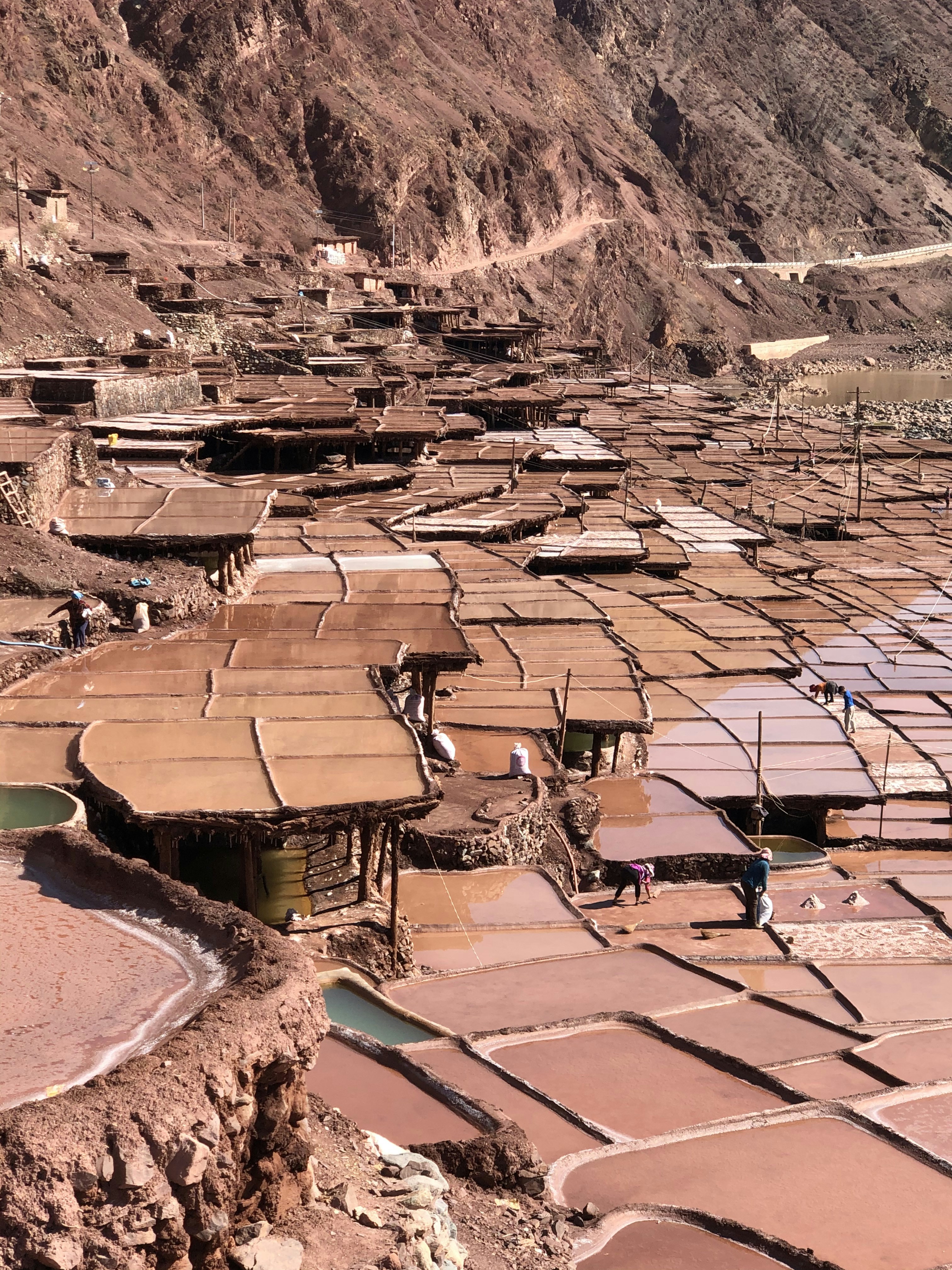 Terraced salt flats in a remote Andean landscape, with workers collecting salt from shallow pools. The intricate patterns highlight traditional harvesting methods.