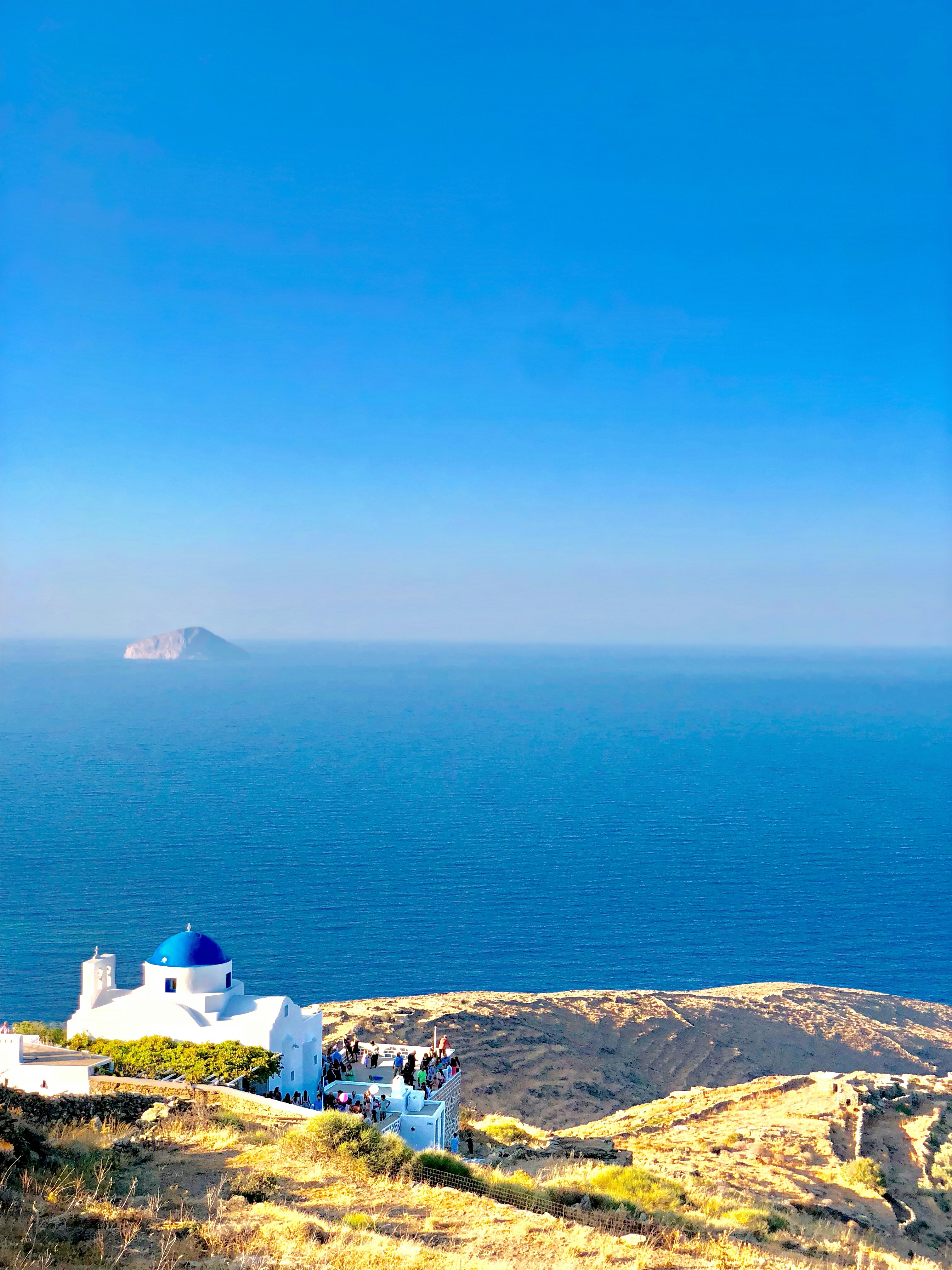 Whitewashed buildings with a blue dome overlook the calm Aegean Sea, with a distant island visible on the horizon.