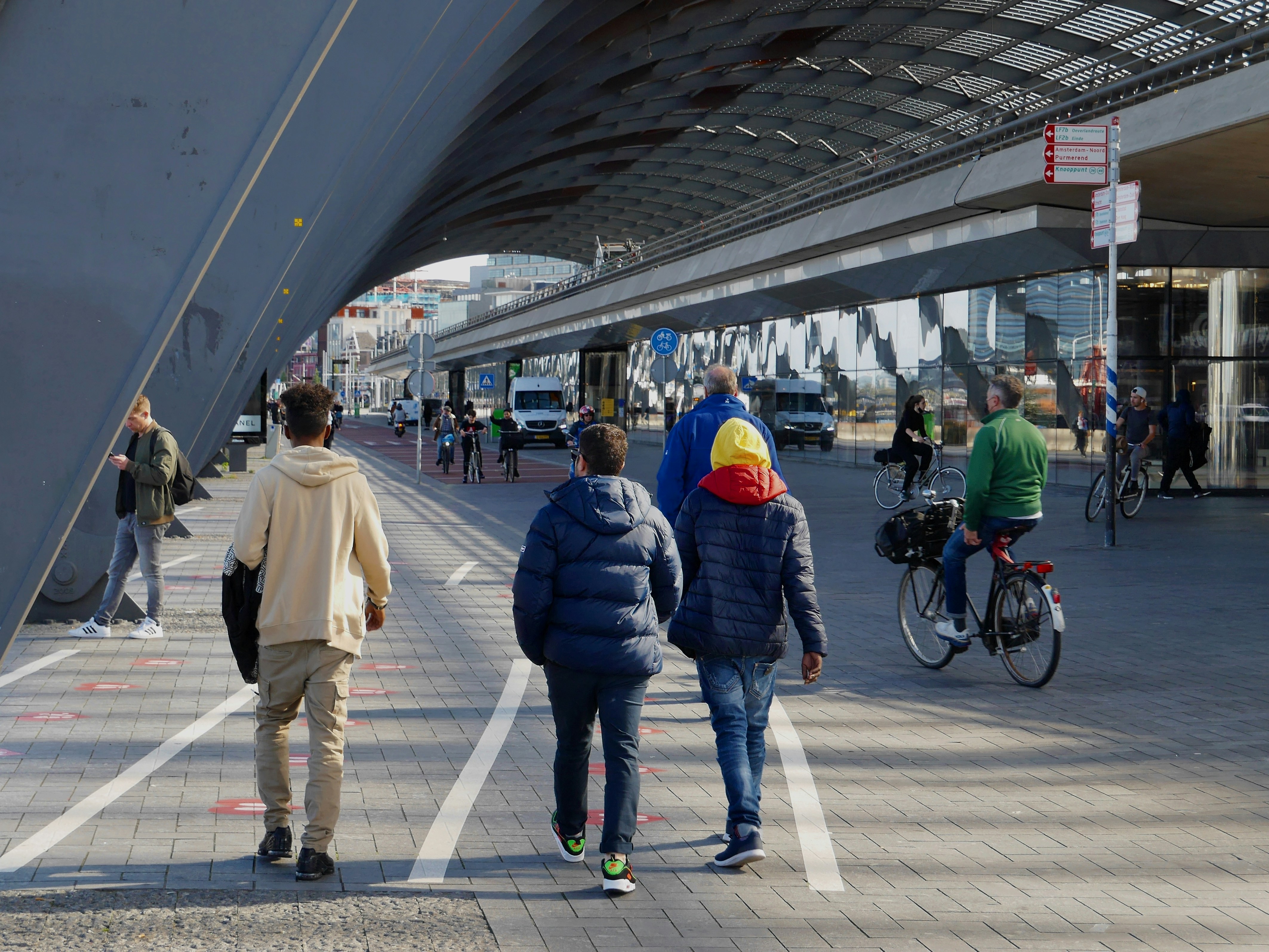 Amsterdam people photo for wallpaper of young urban guys walking together, and biking people under the arched cap of the bus station (above, at the right). This is behind Central Station of Amsterdam city. It is a exclusive area for bikers and pedestrians, just along the border of the IJ river at the left side, with a wide view over the water. Free pic of city photos of Amsterdam, The Netherlands - Dutch human street photography by Fons Heijnsbroek. | people walking on pedestrian lane during daytime