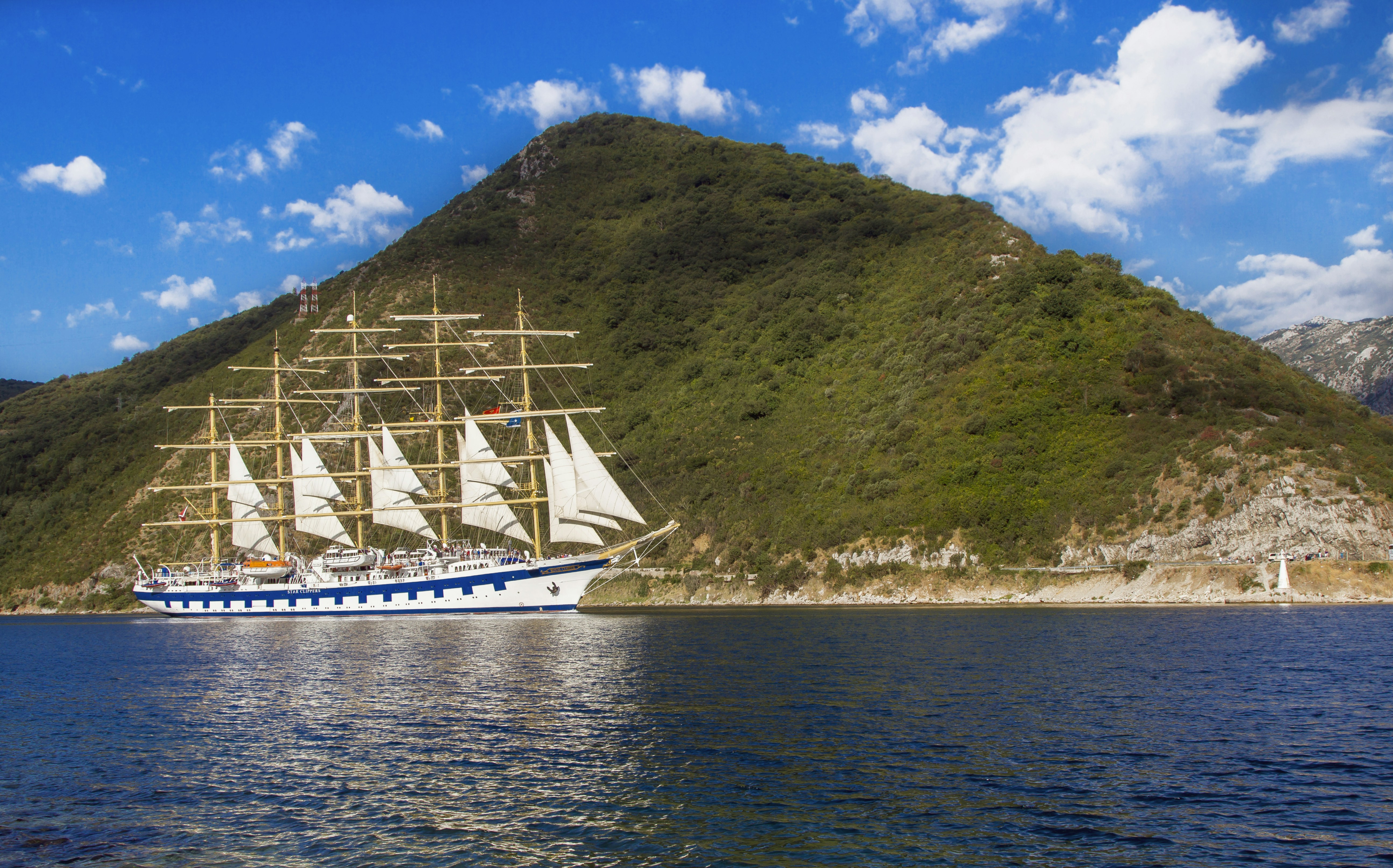Tall ship with white sails gliding across blue water near a lush green mountain under a clear sky.