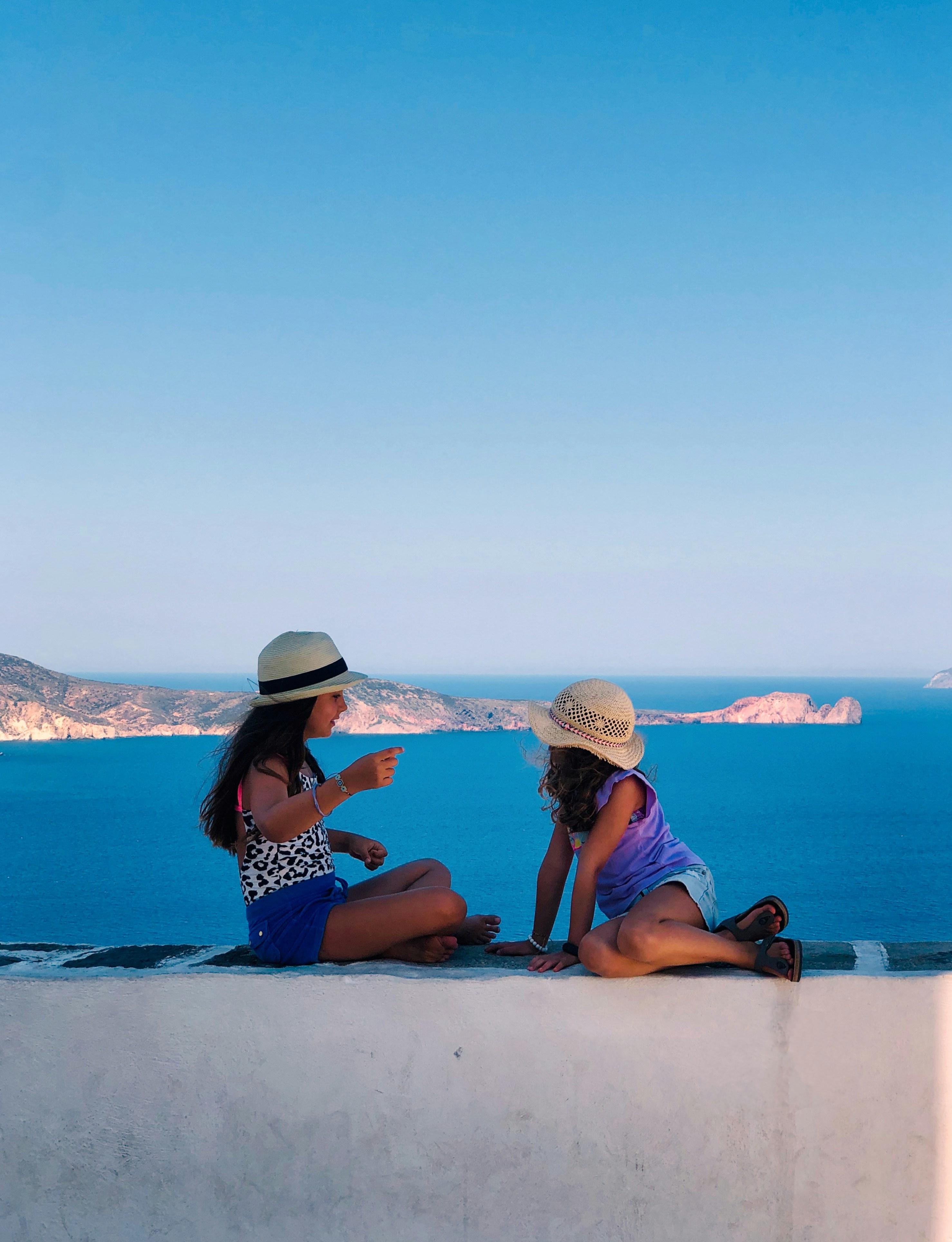 Two girls in summer attire share a moment on a ledge with a stunning ocean view in the background.