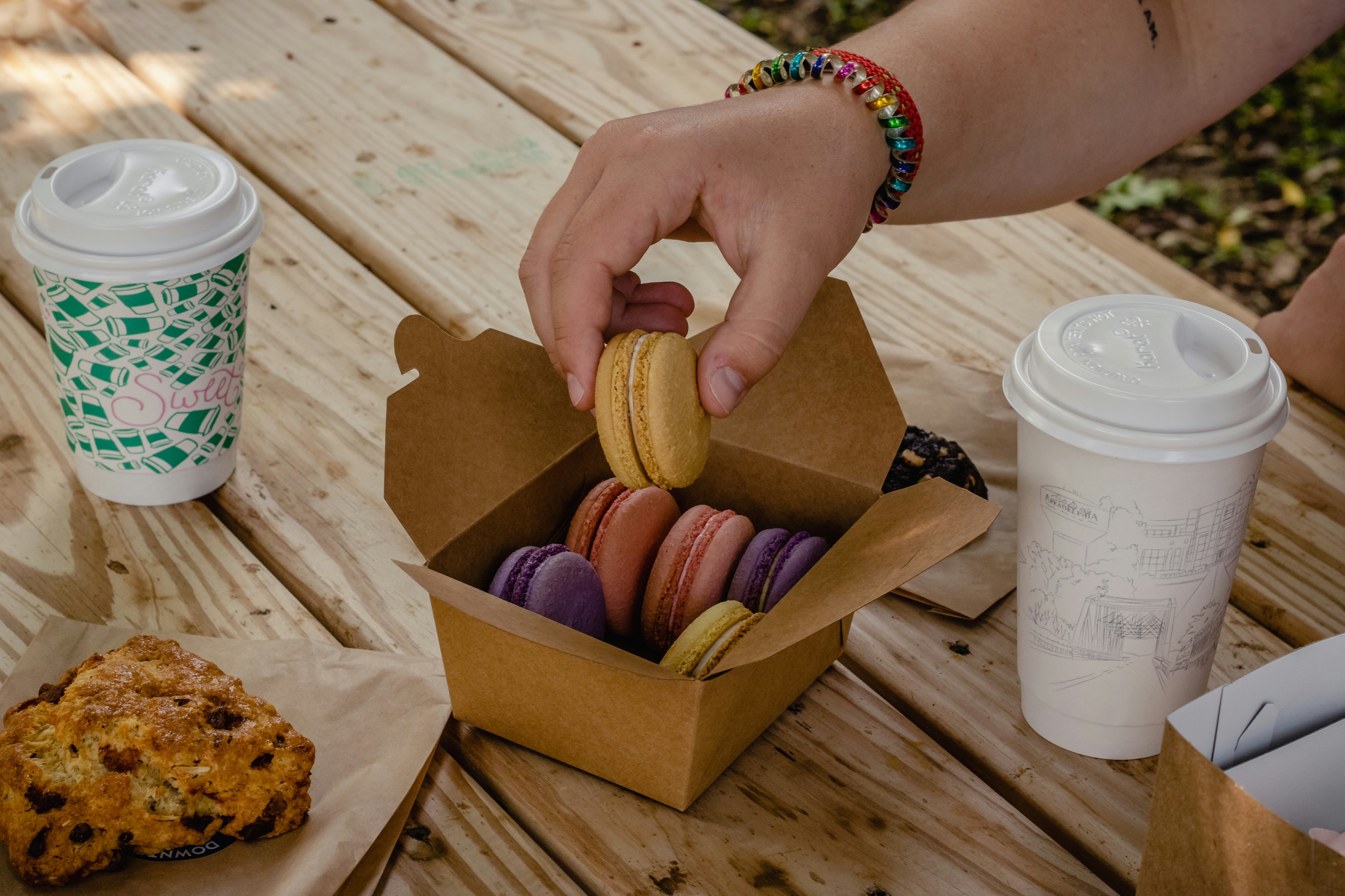 Rainbow Macaron Moment: A Hand Reaches for a Treat atop a Picnic Table
