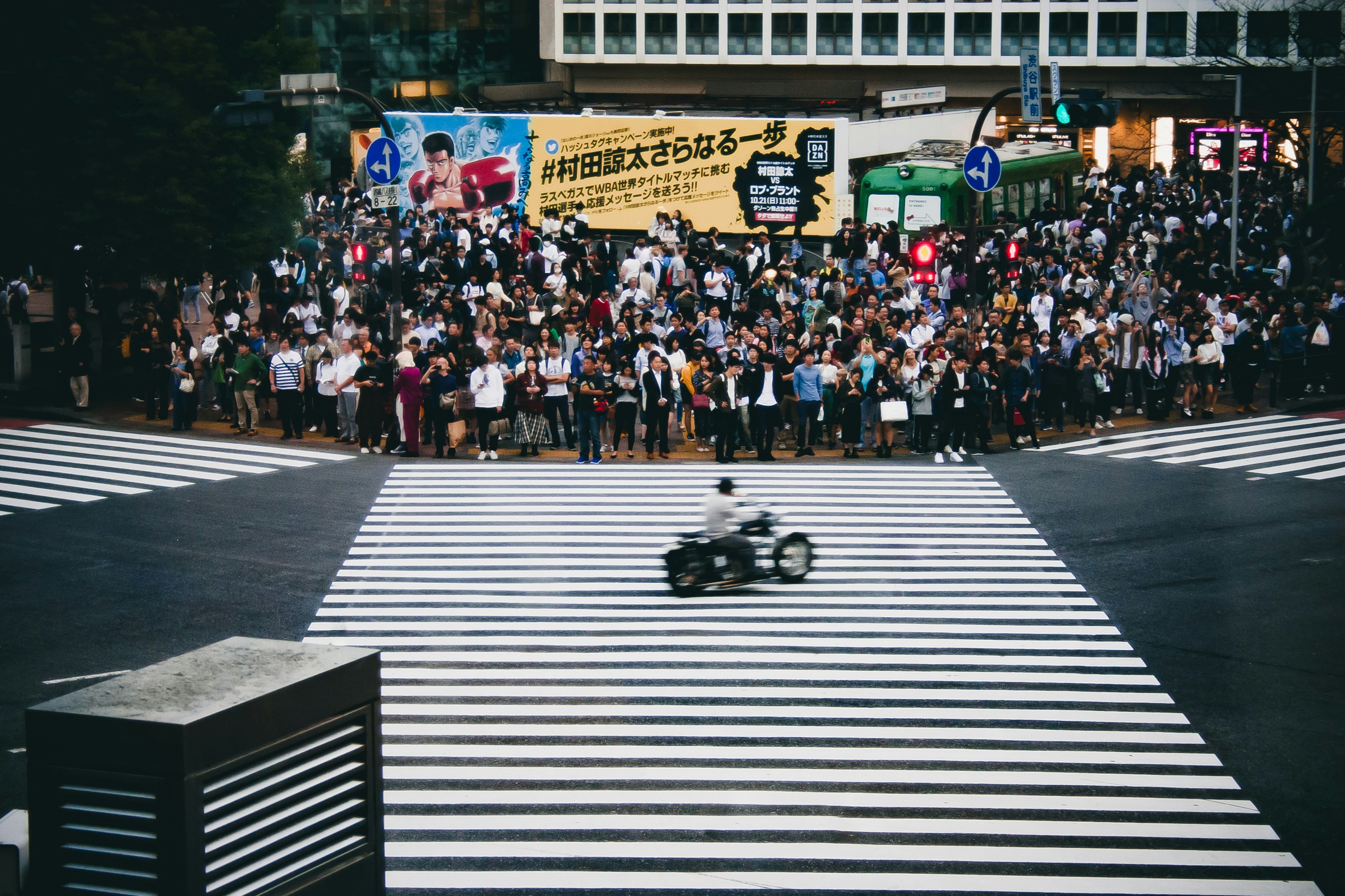 Crowd waits at bustling urban intersection as motorcycle blurs by.