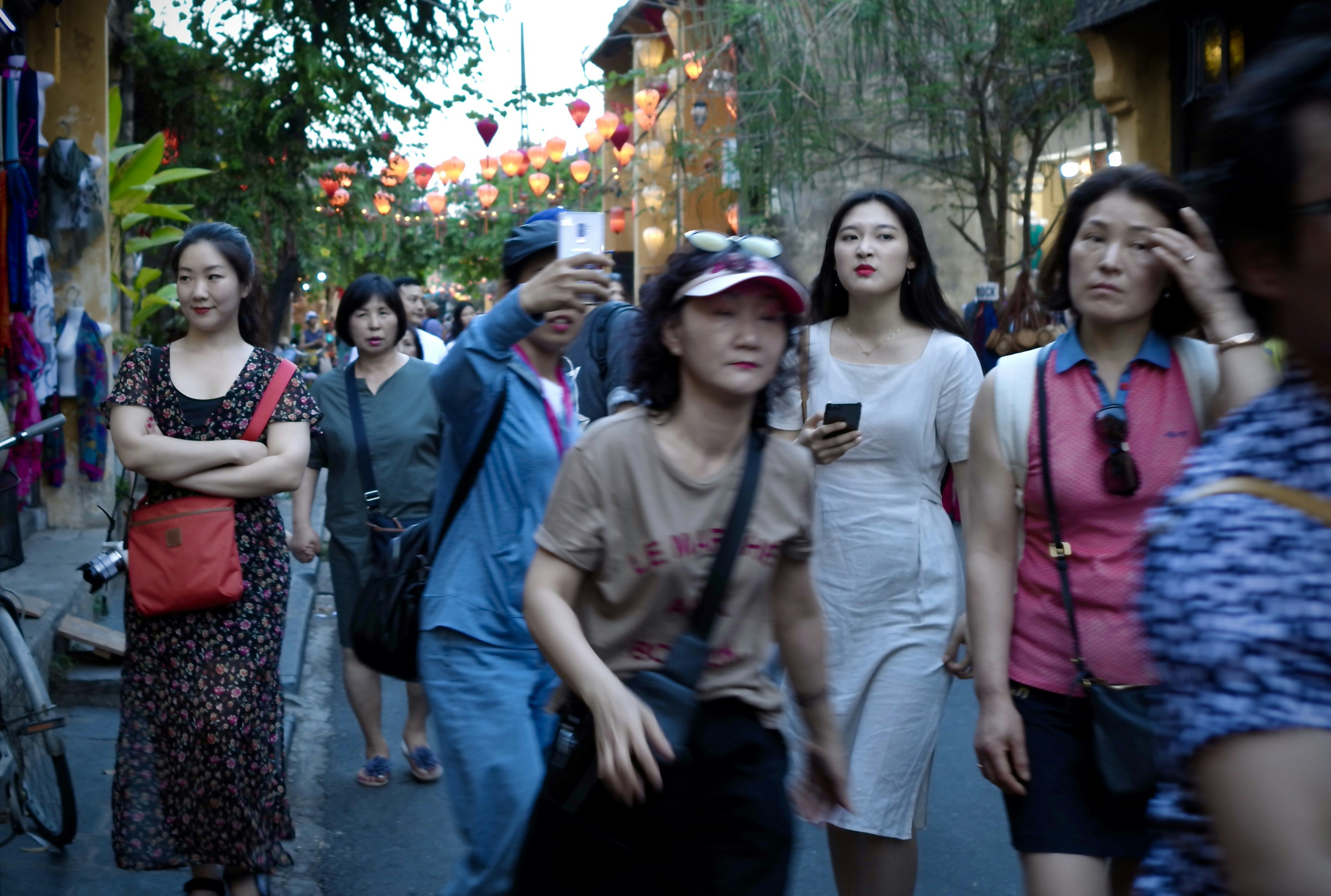 Candid street scene along a lantern-lit avenue; a woman in a white dress stands relatively still as pedestrians move past, capturing a moment of contrast in motion.