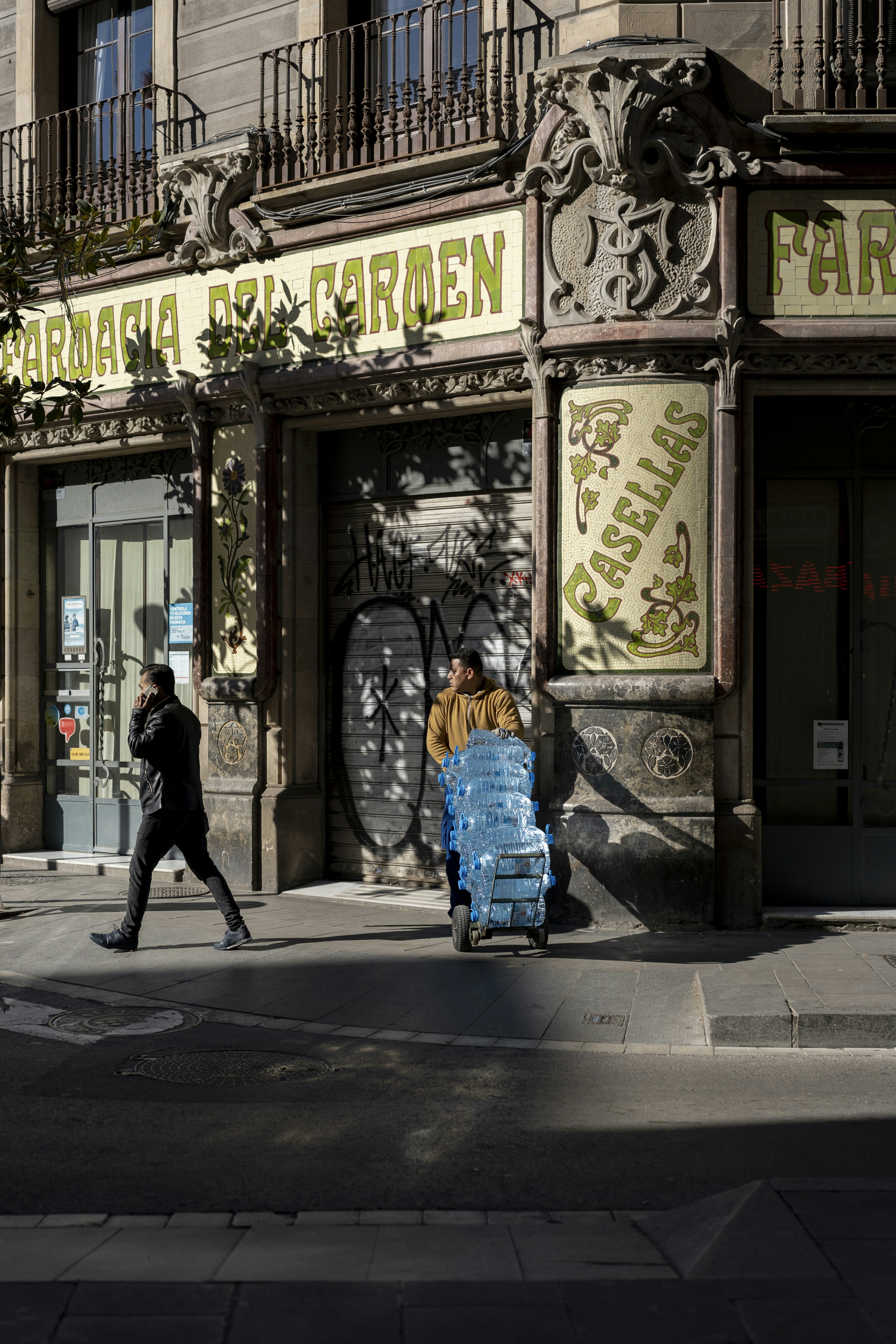 A man stands beside a cart filled with water bottles against a vintage pharmacy facade, capturing the essence of daily life in an urban setting.