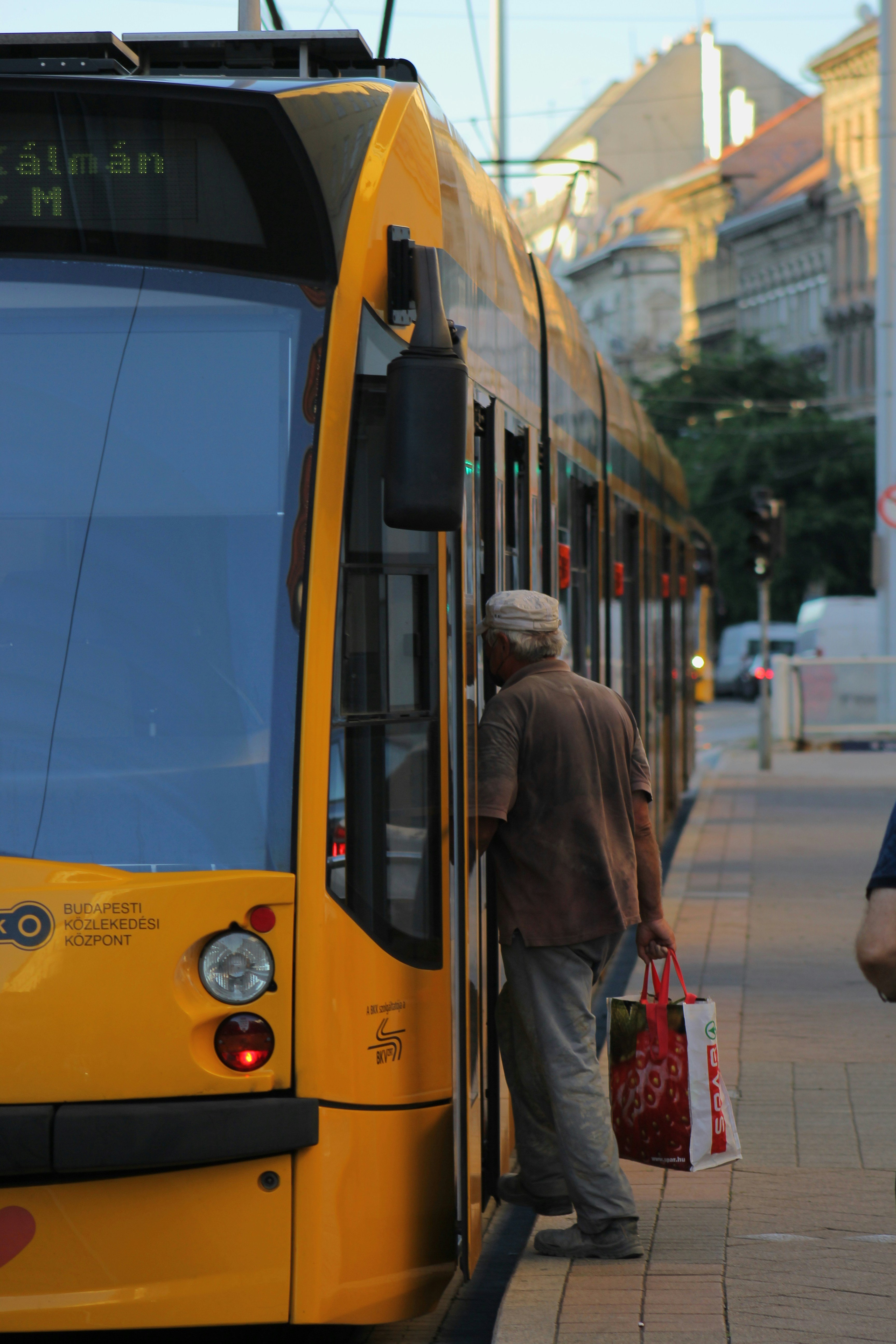 An elderly man boarding a yellow tram, carrying a shopping bag, capturing the essence of daily commuting in an urban setting.