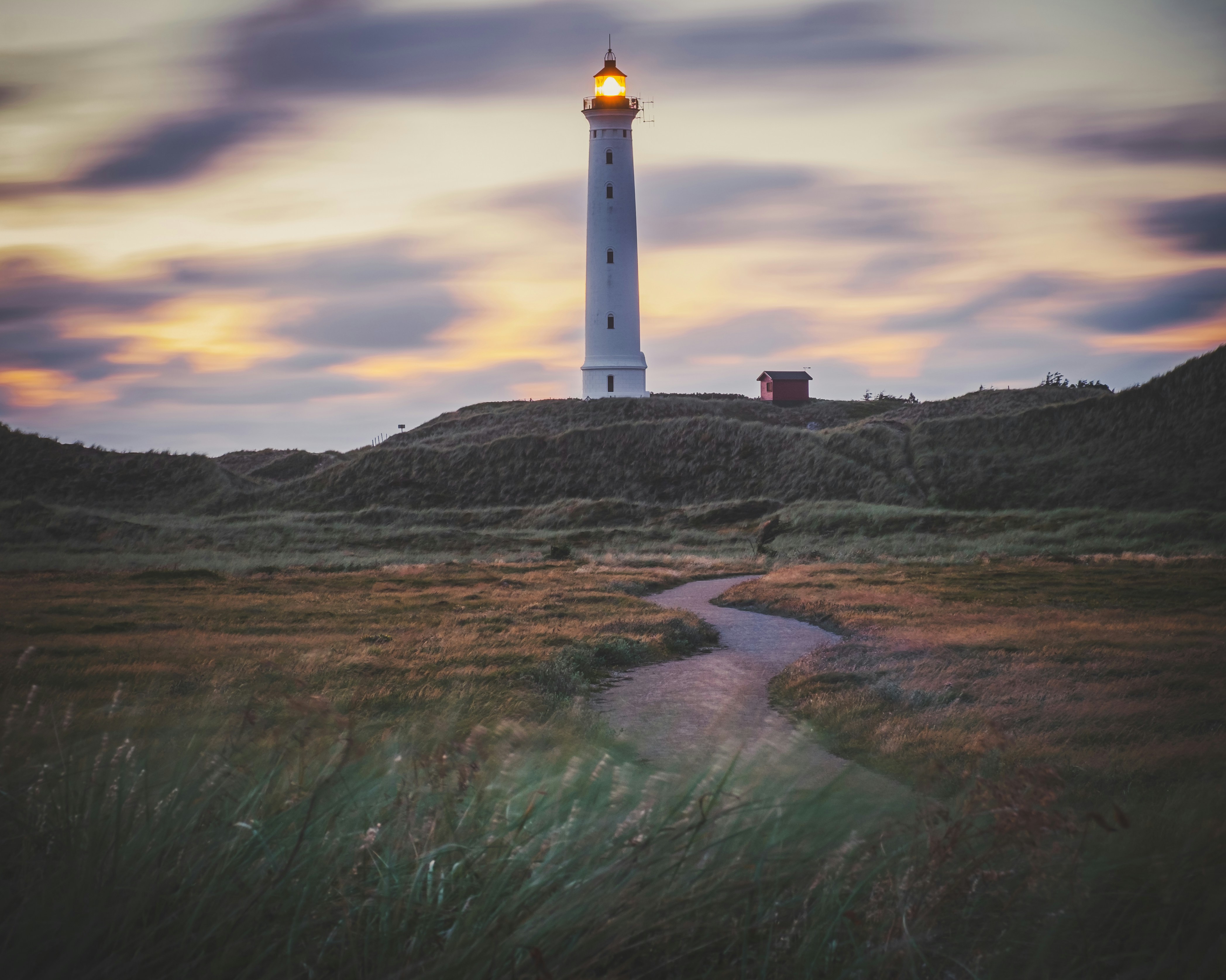 Tall white lighthouse stands on green hills under a dynamic, cloudy sky at sunset.