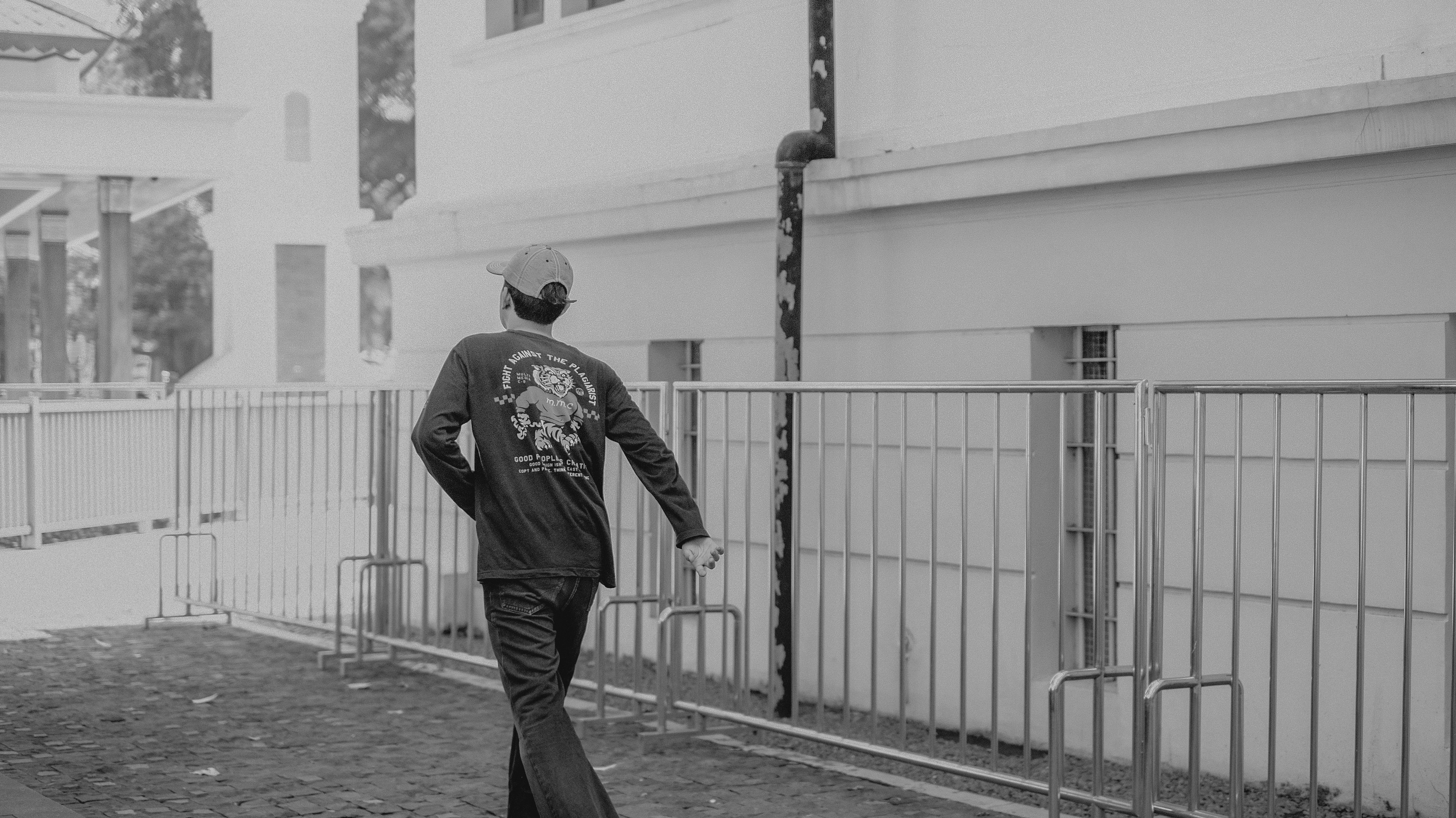 Man walking along a fenced pathway beside a building in black and white.