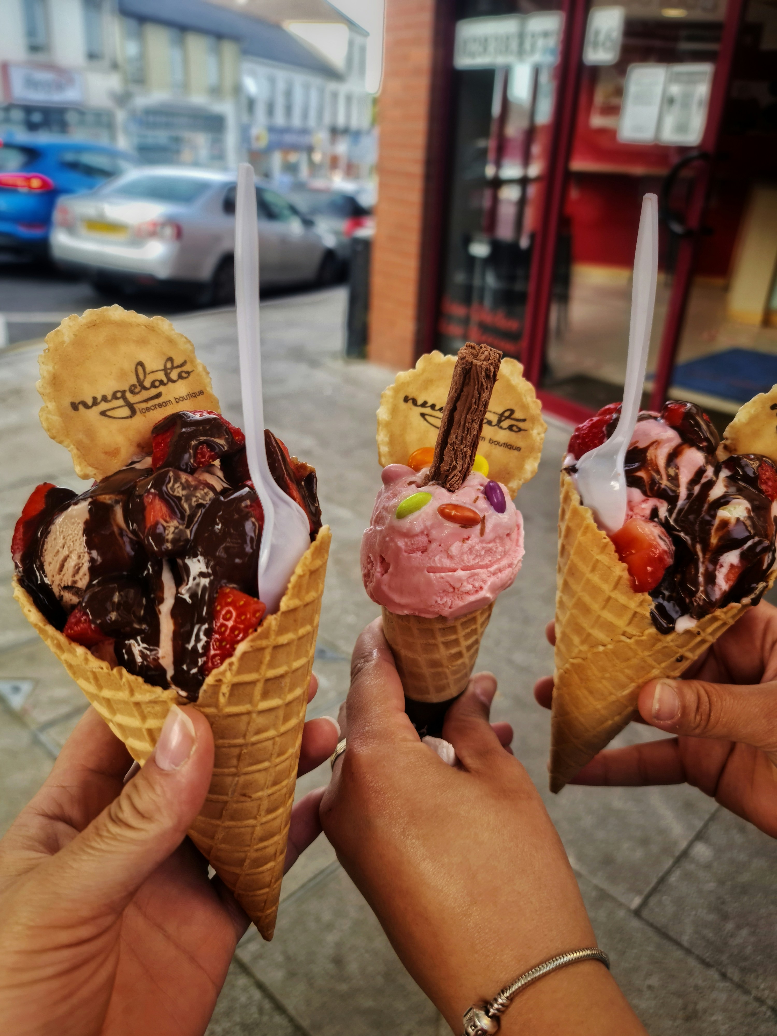 Three hands cradle wafer cones—chocolate, pink strawberry, and a topped variant. A city street with storefronts and parked cars blurs in the background.