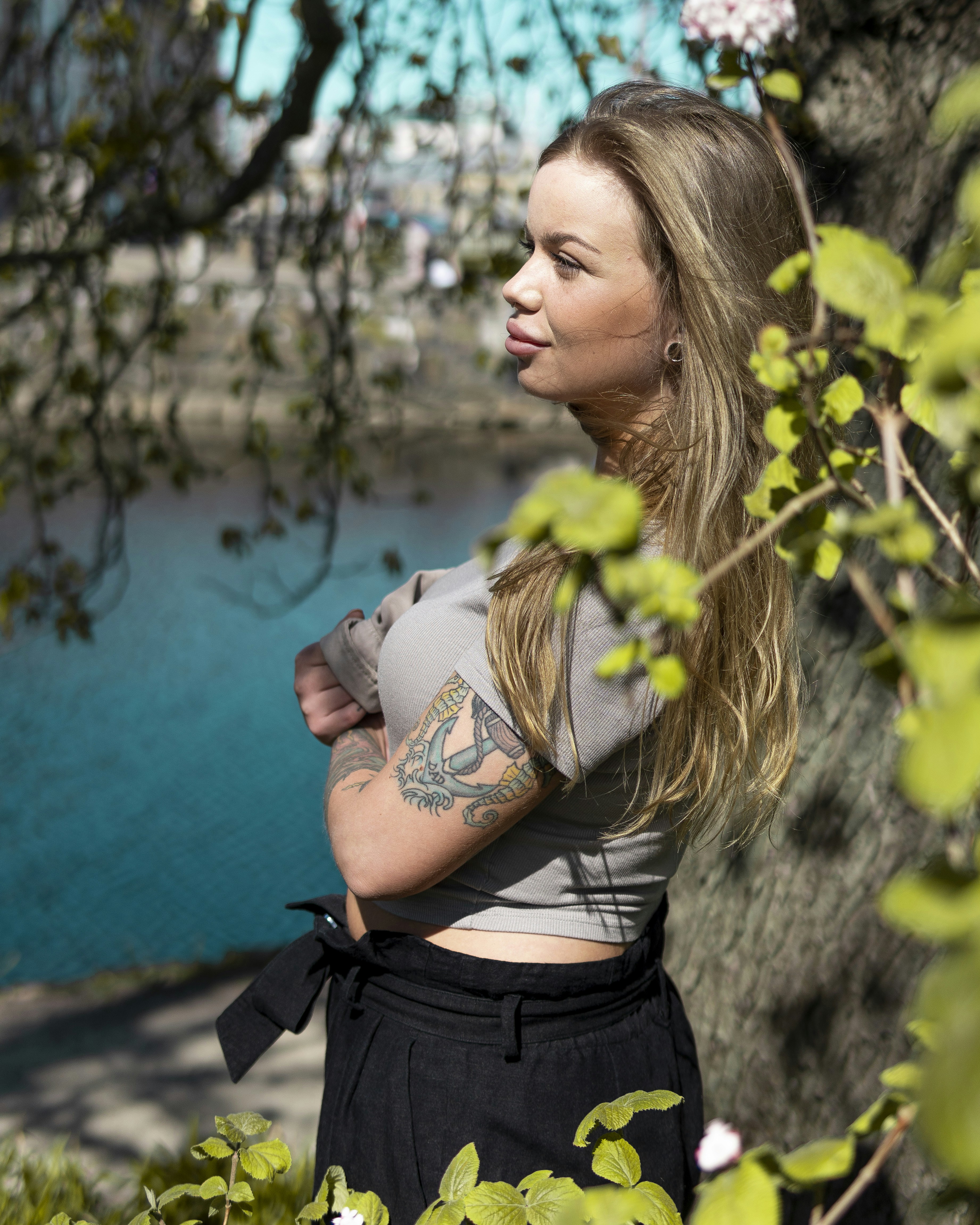 A young woman gazes thoughtfully while framed by vibrant green foliage, with a tranquil blue water backdrop. Her tattoo and relaxed pose add character to the scene.