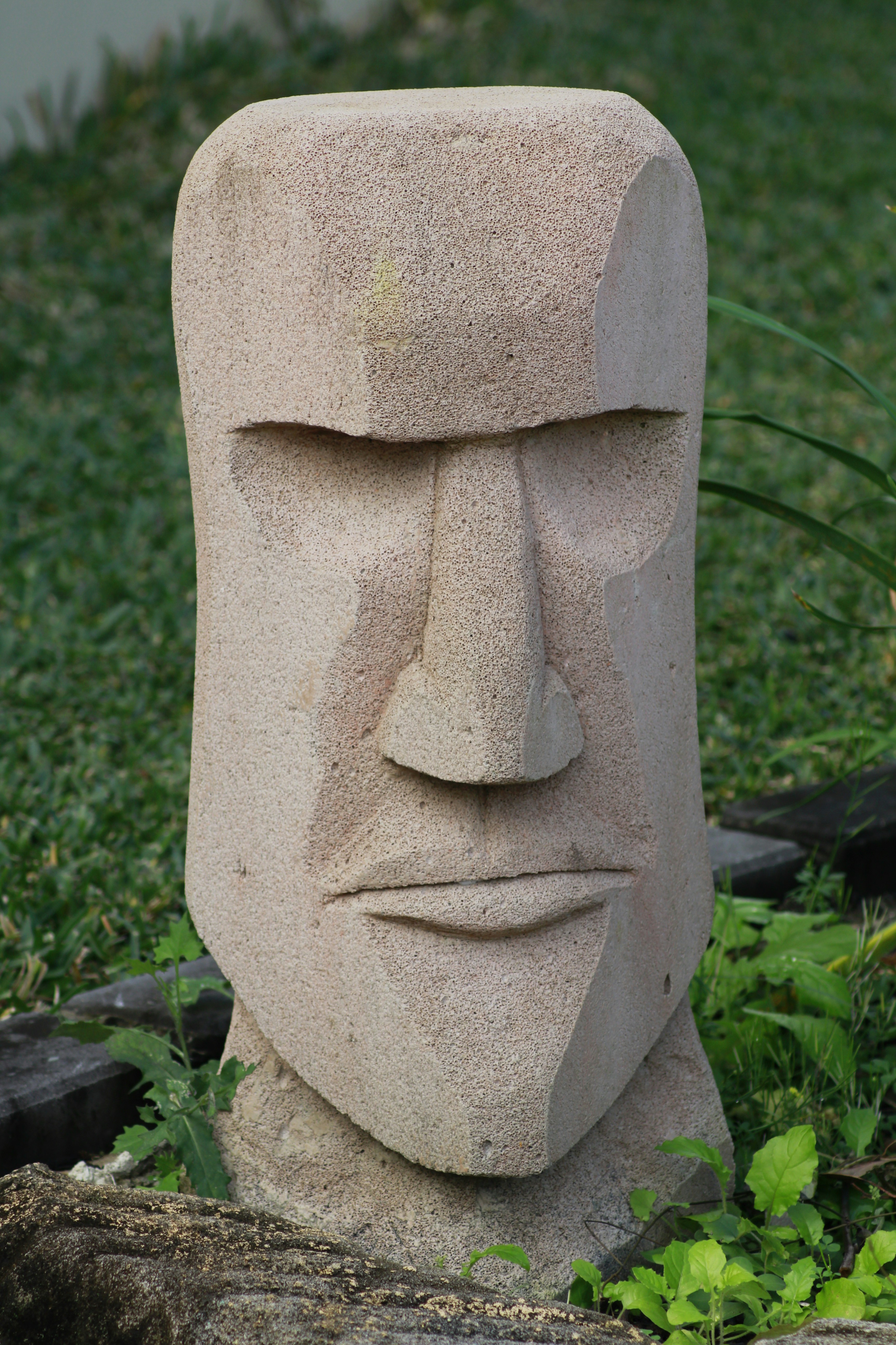 A carved stone statue resembling a Moai head, set against a backdrop of green grass and foliage.
