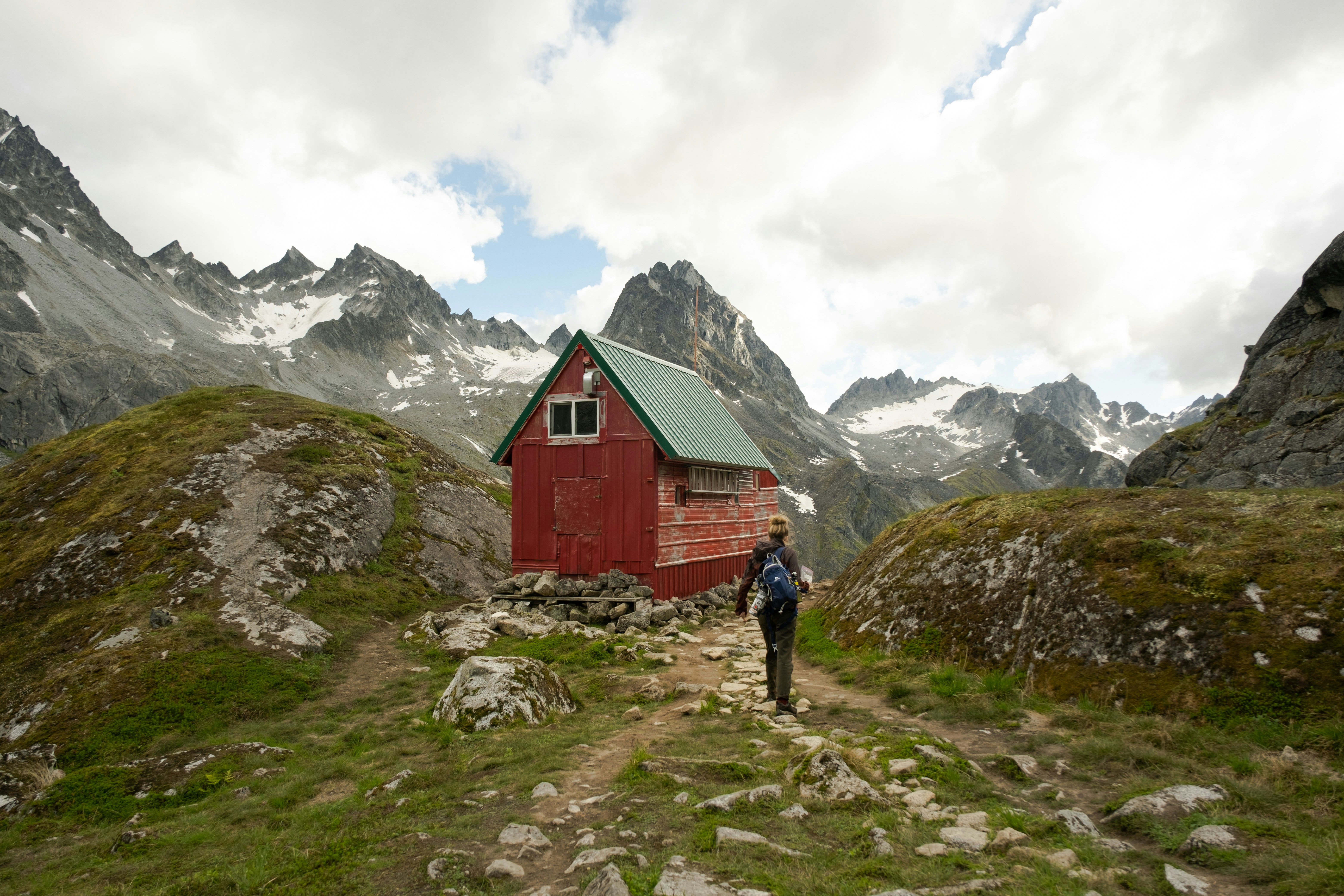 Red Alpine Cabin Anchoring a Rugged Trail Under Snow-Capped Peaks